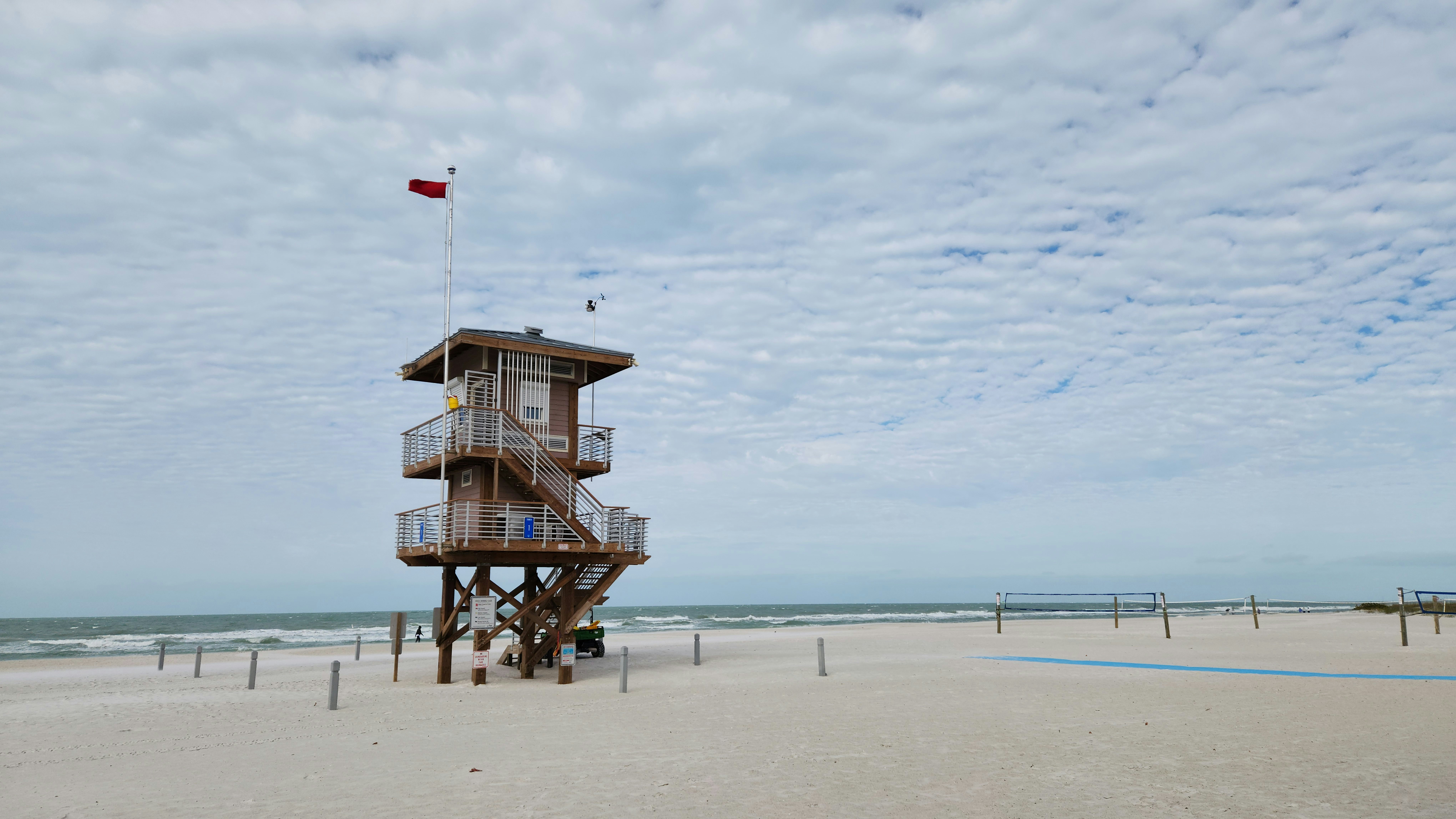 This photograph captures a wooden lifeguard tower standing tall on a sandy beach. A red flag, indicating caution due to hazardous conditions, is flying atop the tower. The sky above is partly cloudy with a pattern of altocumulus clouds, and the ocean in the background appears choppy. The beach is nearly empty, adding to the sense of caution suggested by the red flag.