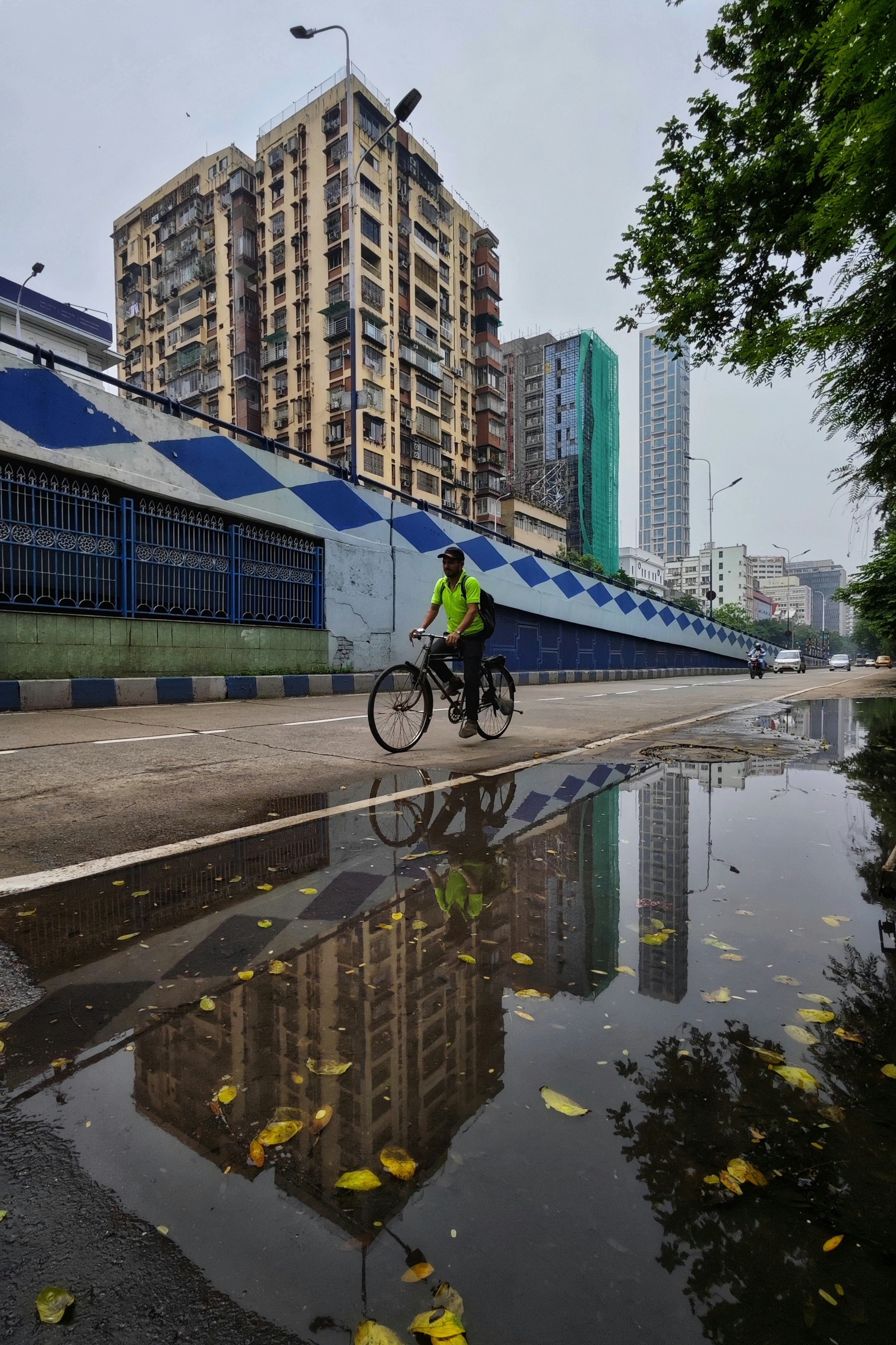 Bright-green cyclist rides along a rain-soaked urban street, with a large puddle reflecting the surrounding buildings and blue fencing.