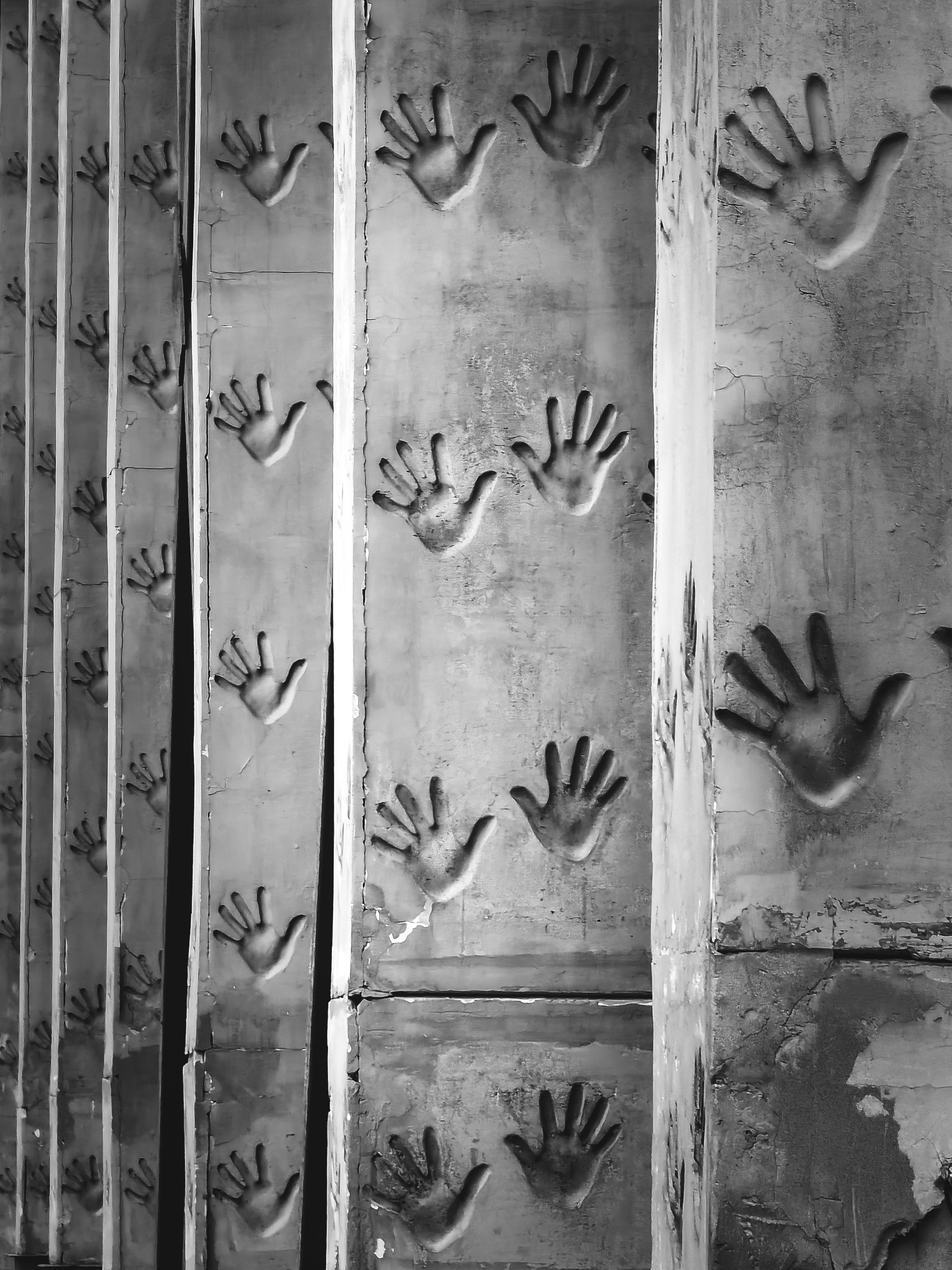 Monochrome wall of weathered concrete panels with numerous raised handprints arranged in vertical strips, creating a repetitive pattern.