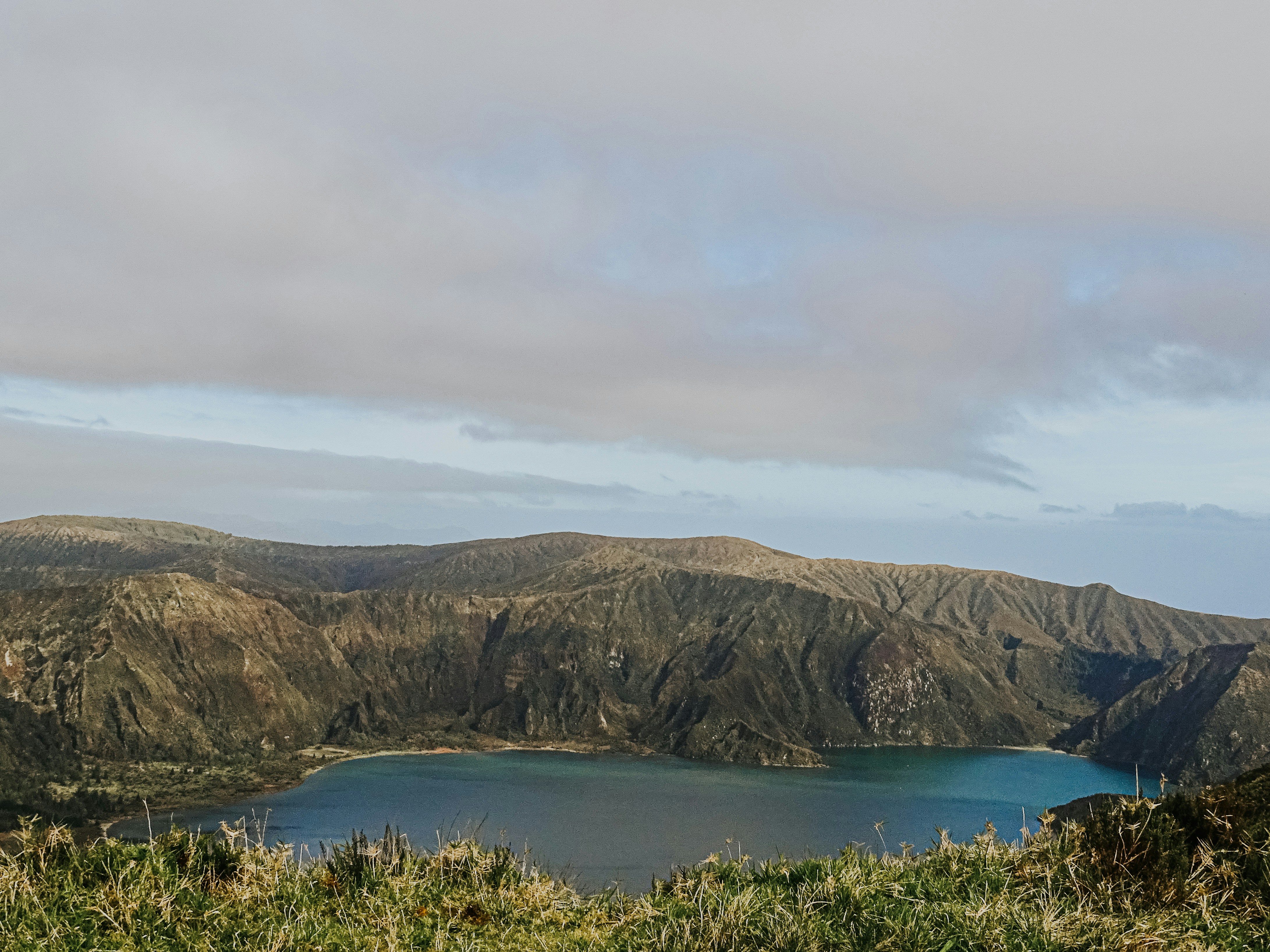 Caldera lake framed by rugged cliffs under a pale sky. Foreground shrubs anchor the scene, highlighting turquoise water and the dramatic shoreline.