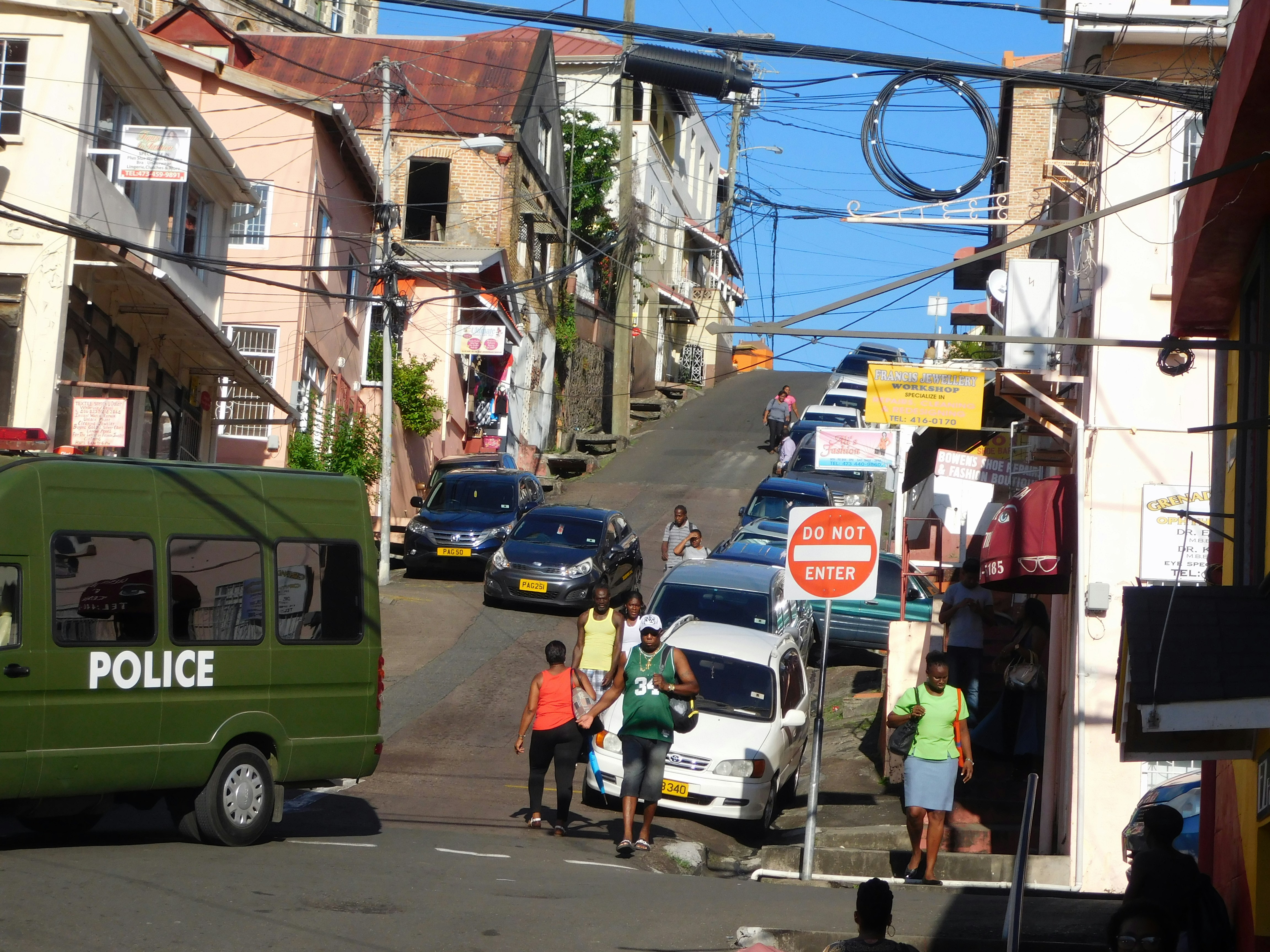 A bustling street scene in a Caribbean town, showcasing local pedestrians and vehicles, with a police van prominently parked. The vibrant atmosphere reflects daily life in the community.