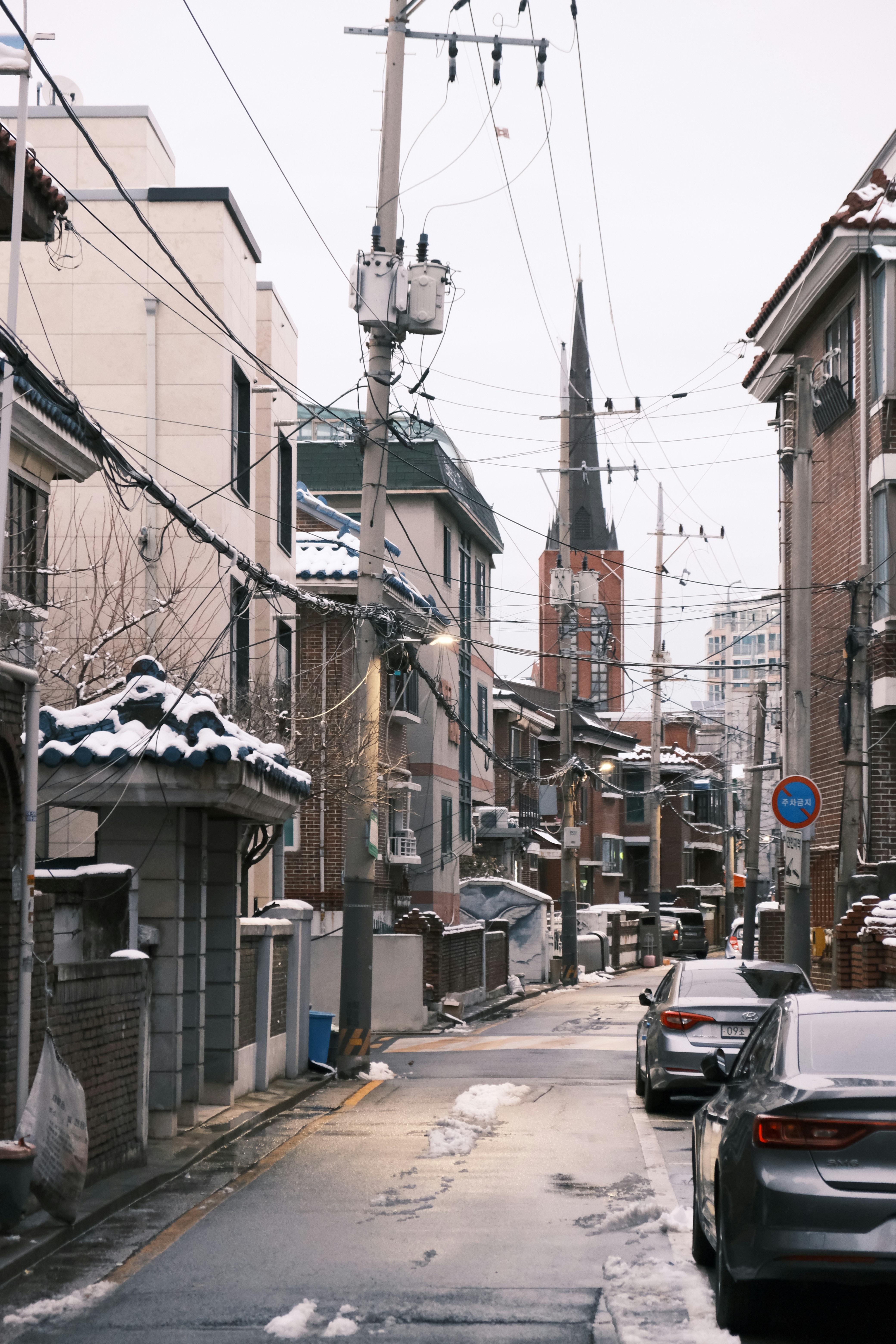 Snow-dusted urban street lined with parked cars and traditional rooftops, leading to a distant church spire.
