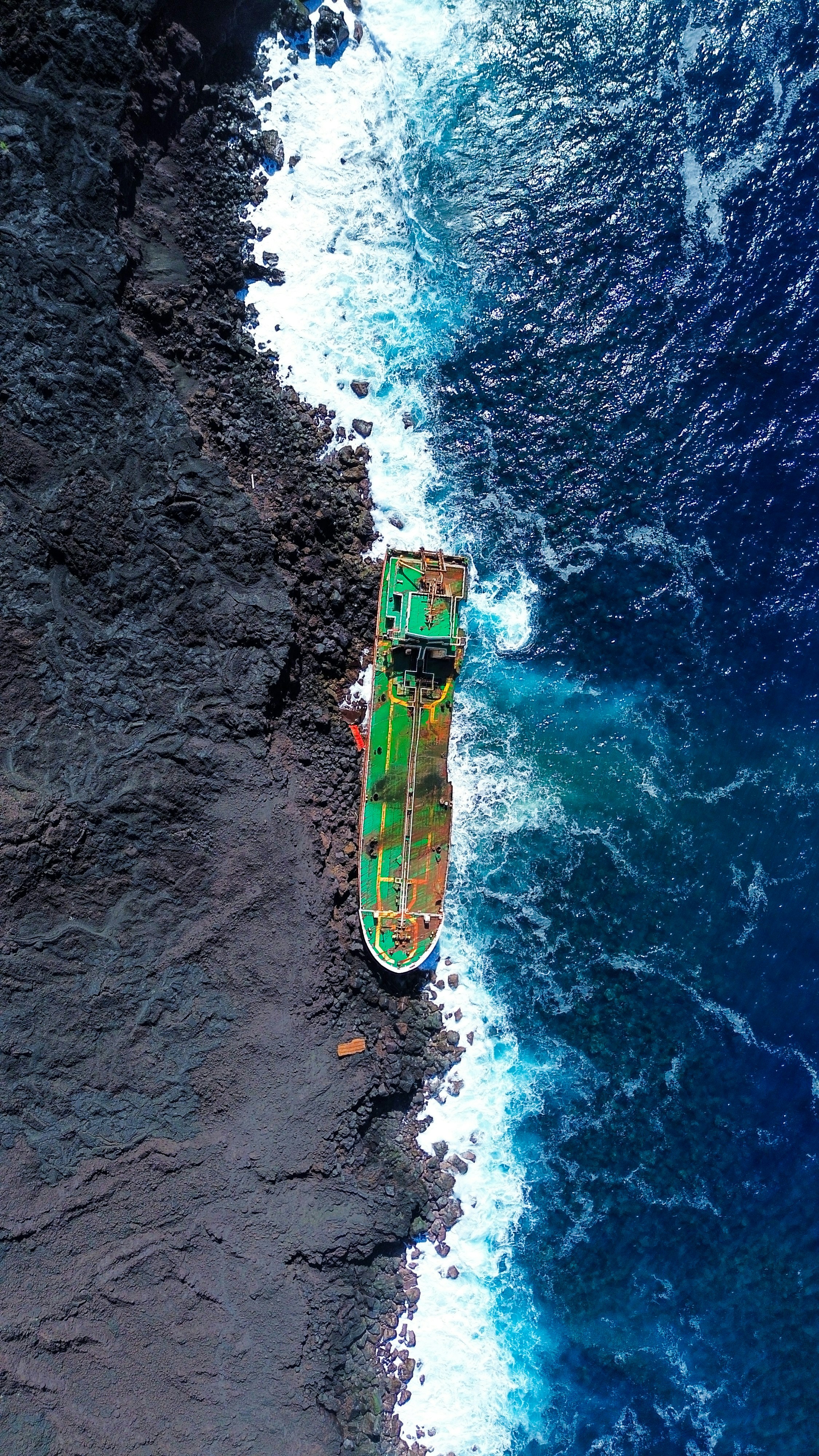 Drone shot of a weathered green workboat wedged along a lava-black coastline with surf crashing on jagged rocks and deep blue water beyond.