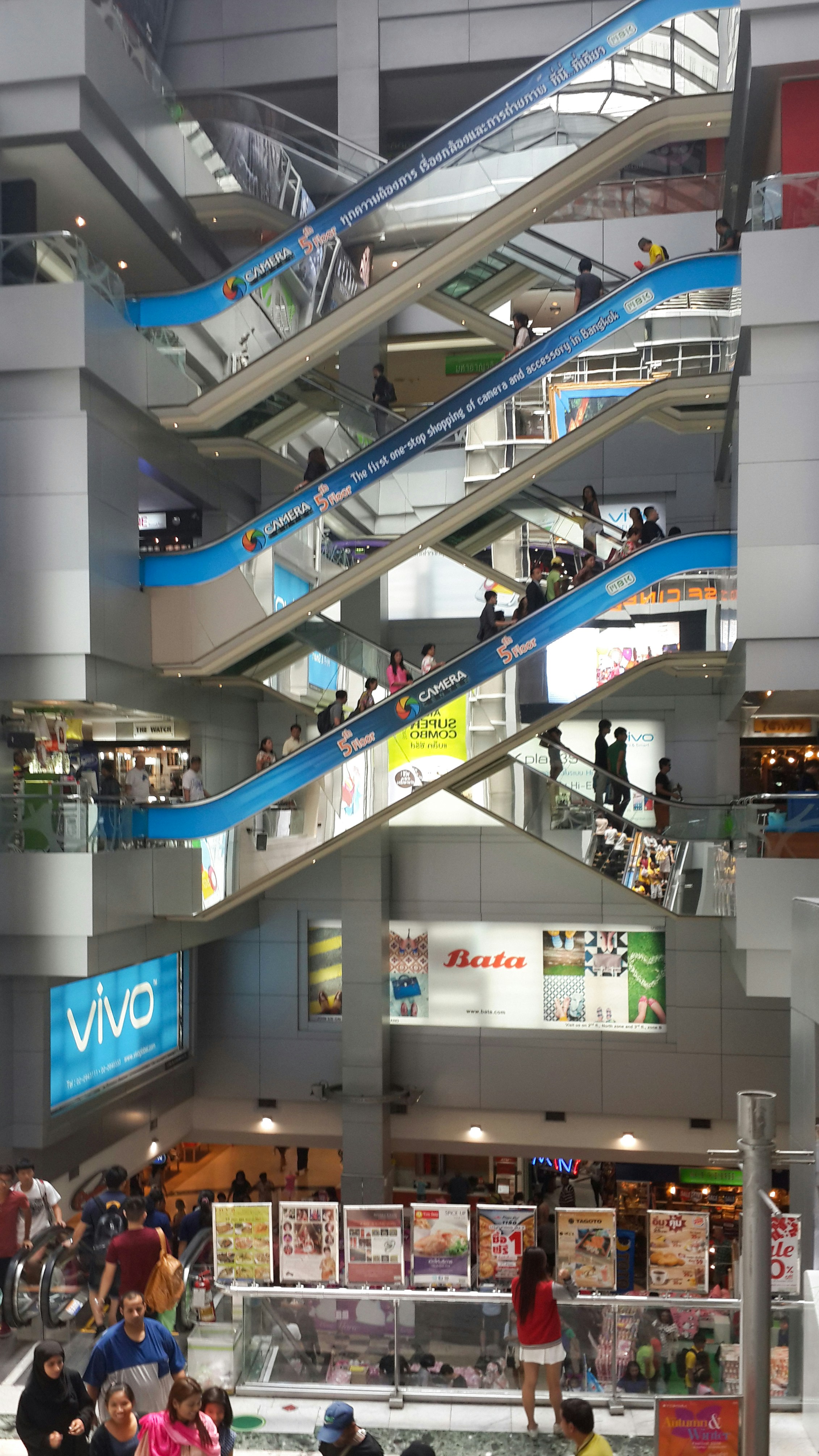 A multi-level shopping center showcasing escalators filled with shoppers, vibrant advertisements, and a bustling atmosphere.