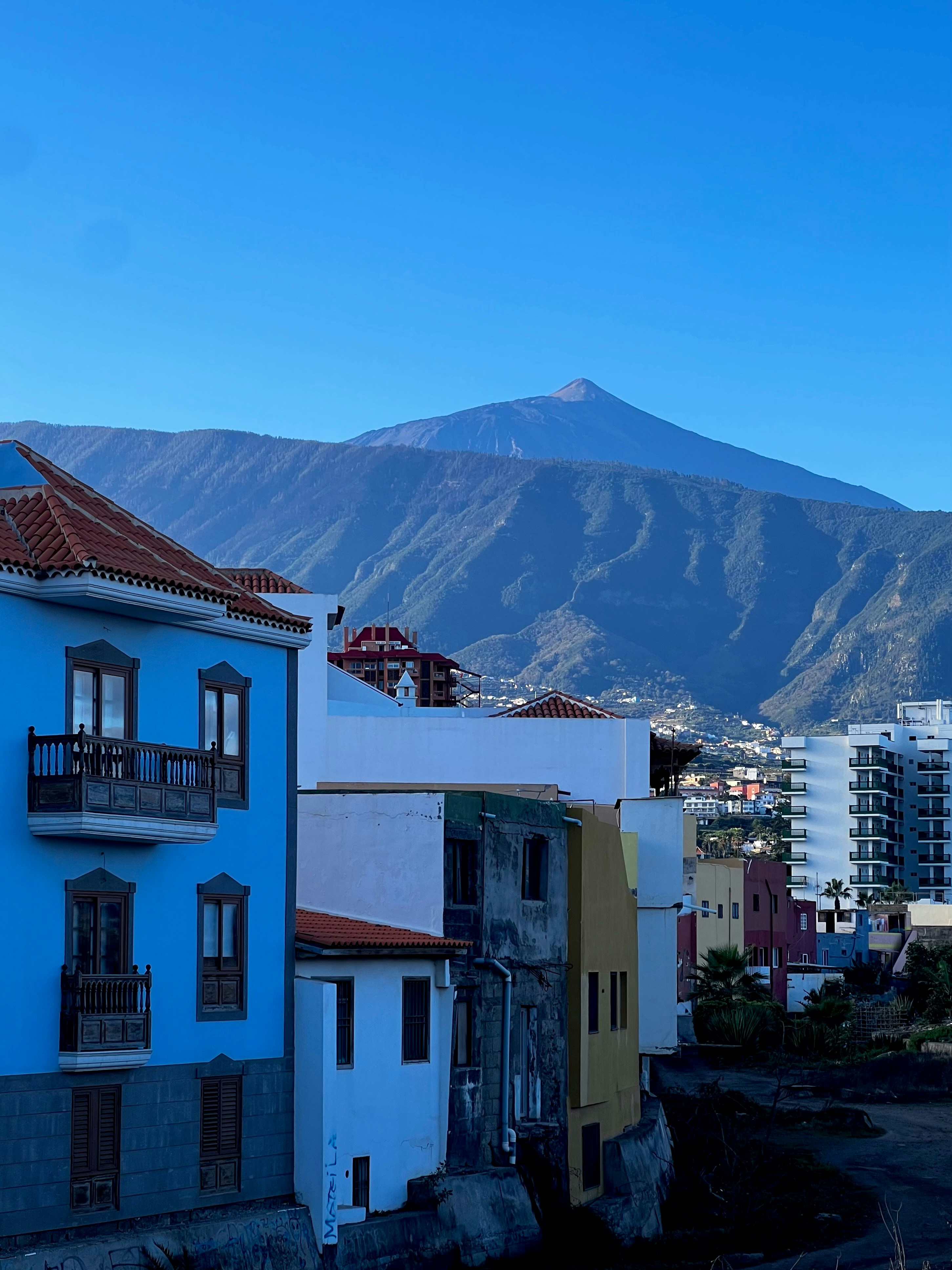 Colorful buildings line the waterfront, with a majestic mountain rising in the background under a clear blue sky.