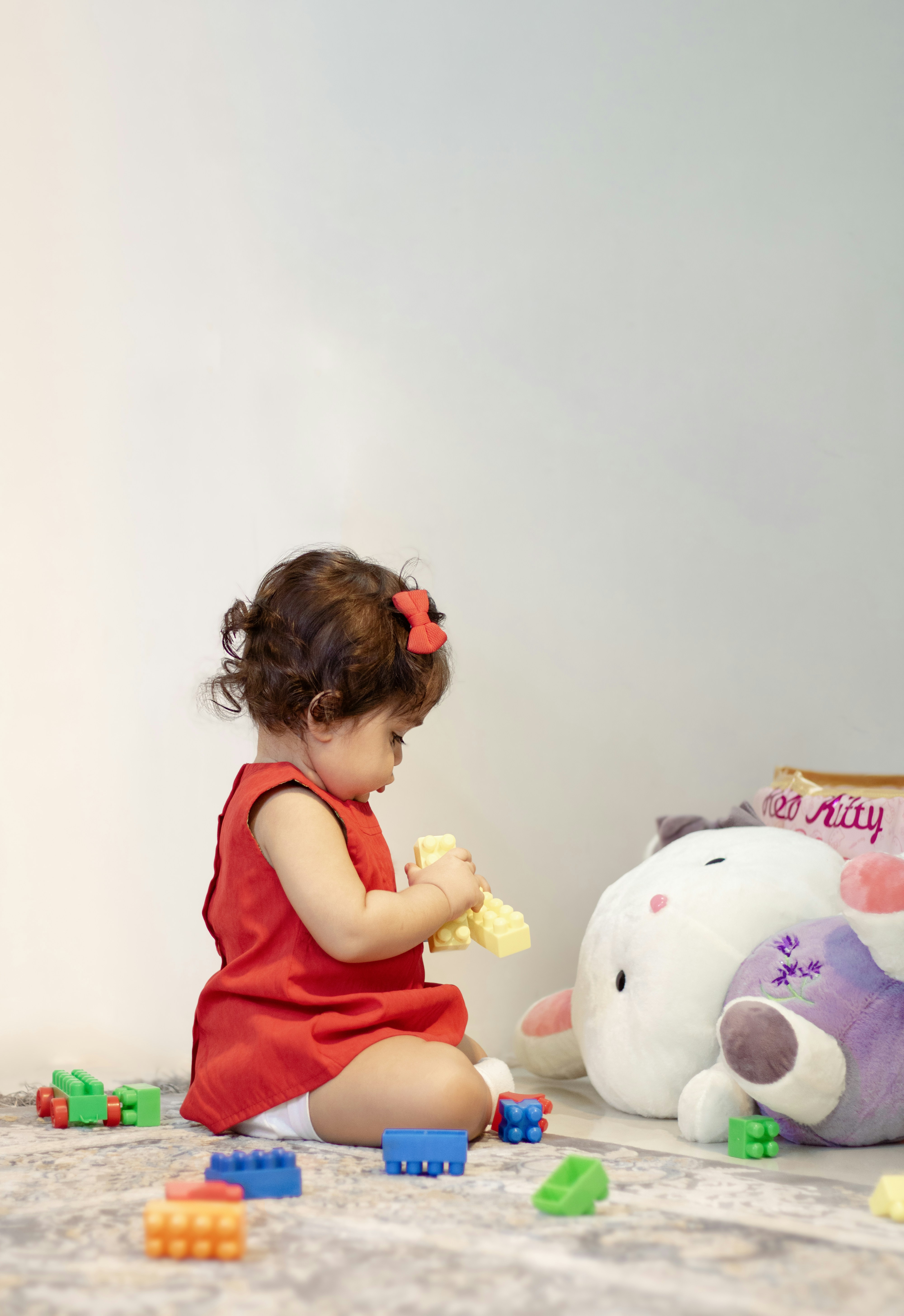 Toddler in a red dress assembling colorful building blocks beside a plush toy. The soft, neutral background highlights the playful scene.