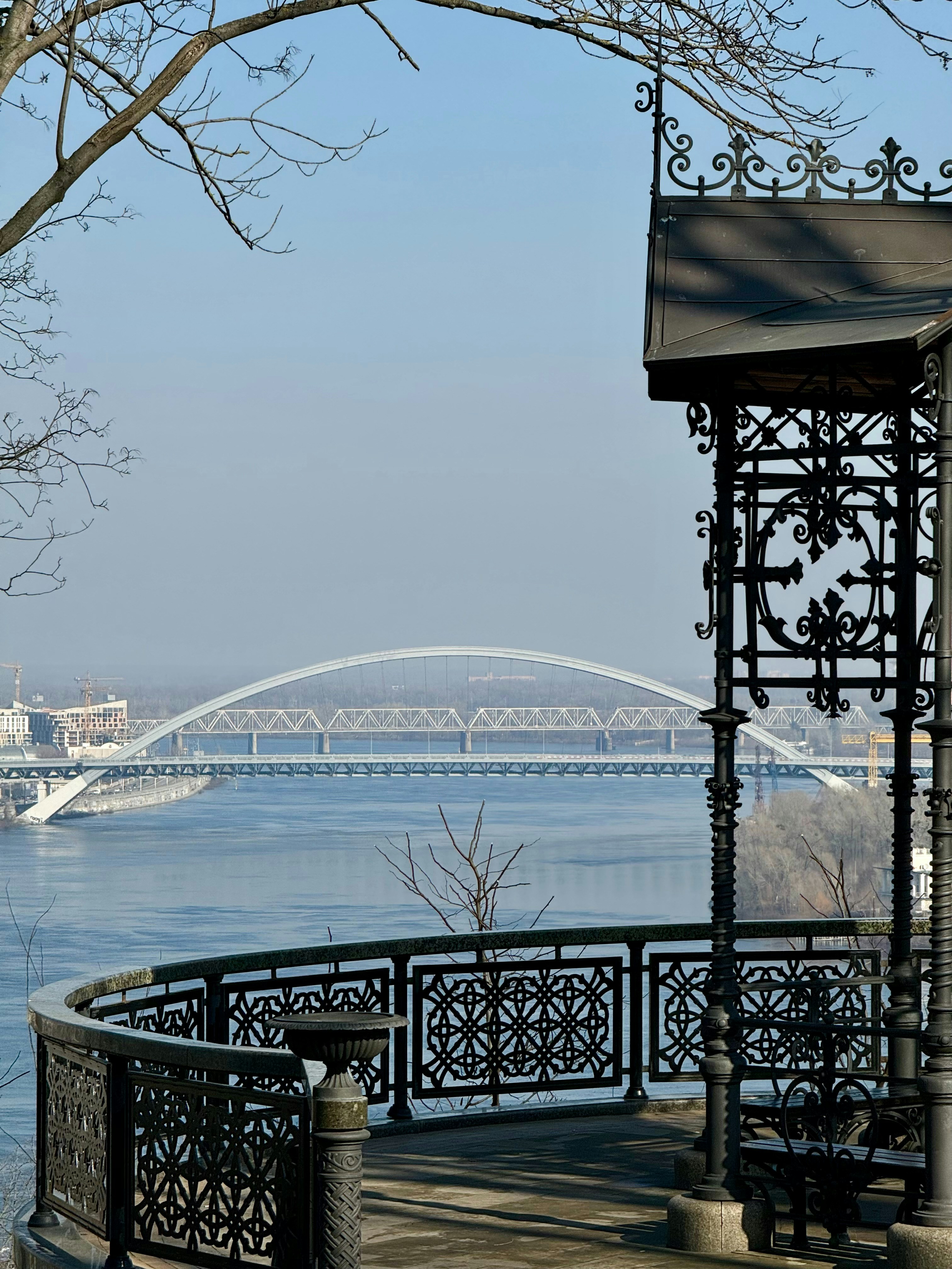 Ornate gazebo overlooking a serene river with a modern bridge in the background, framed by delicate branches. 