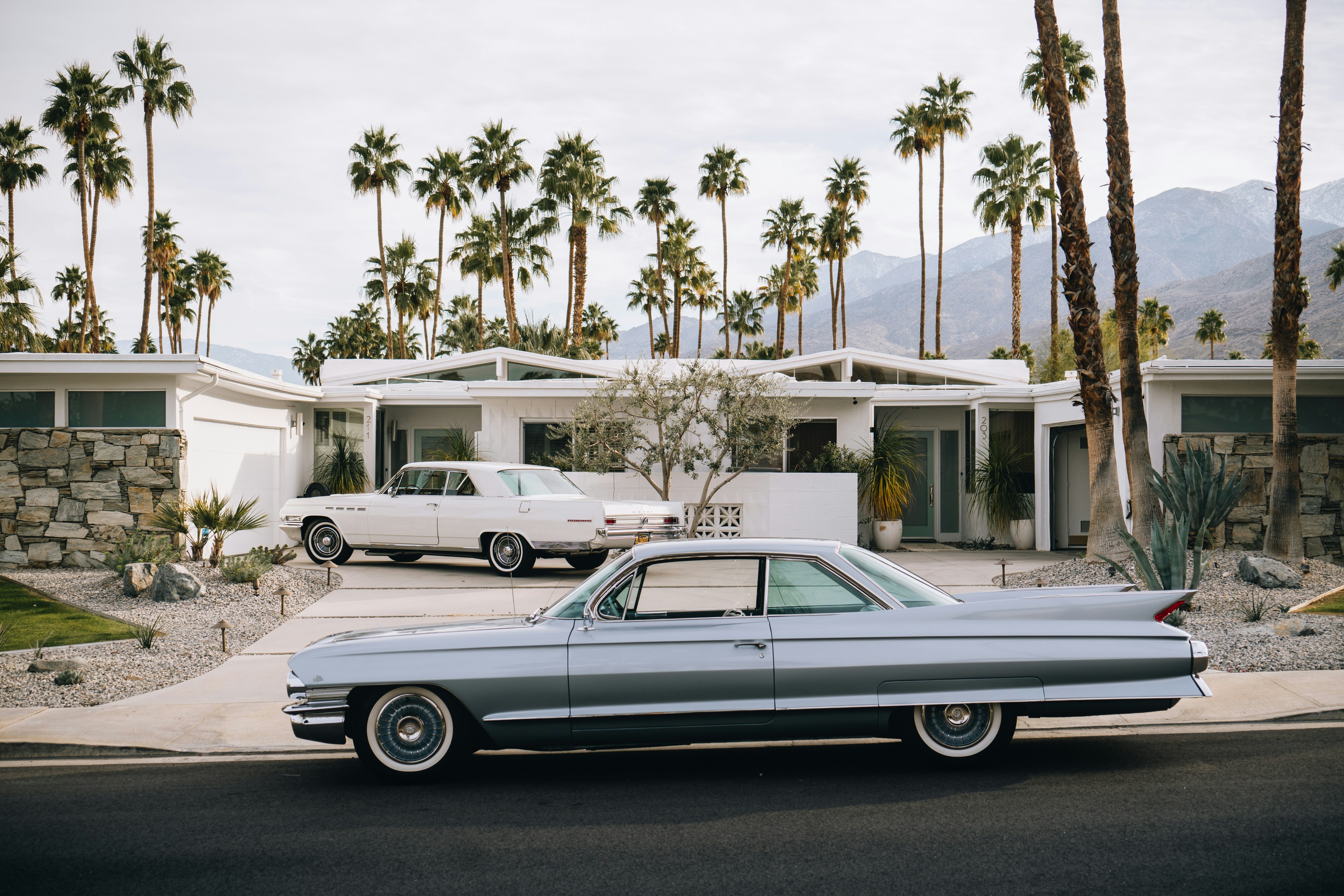 Classic cars parked outside a mid-century modern house with palm trees and mountains in the background.