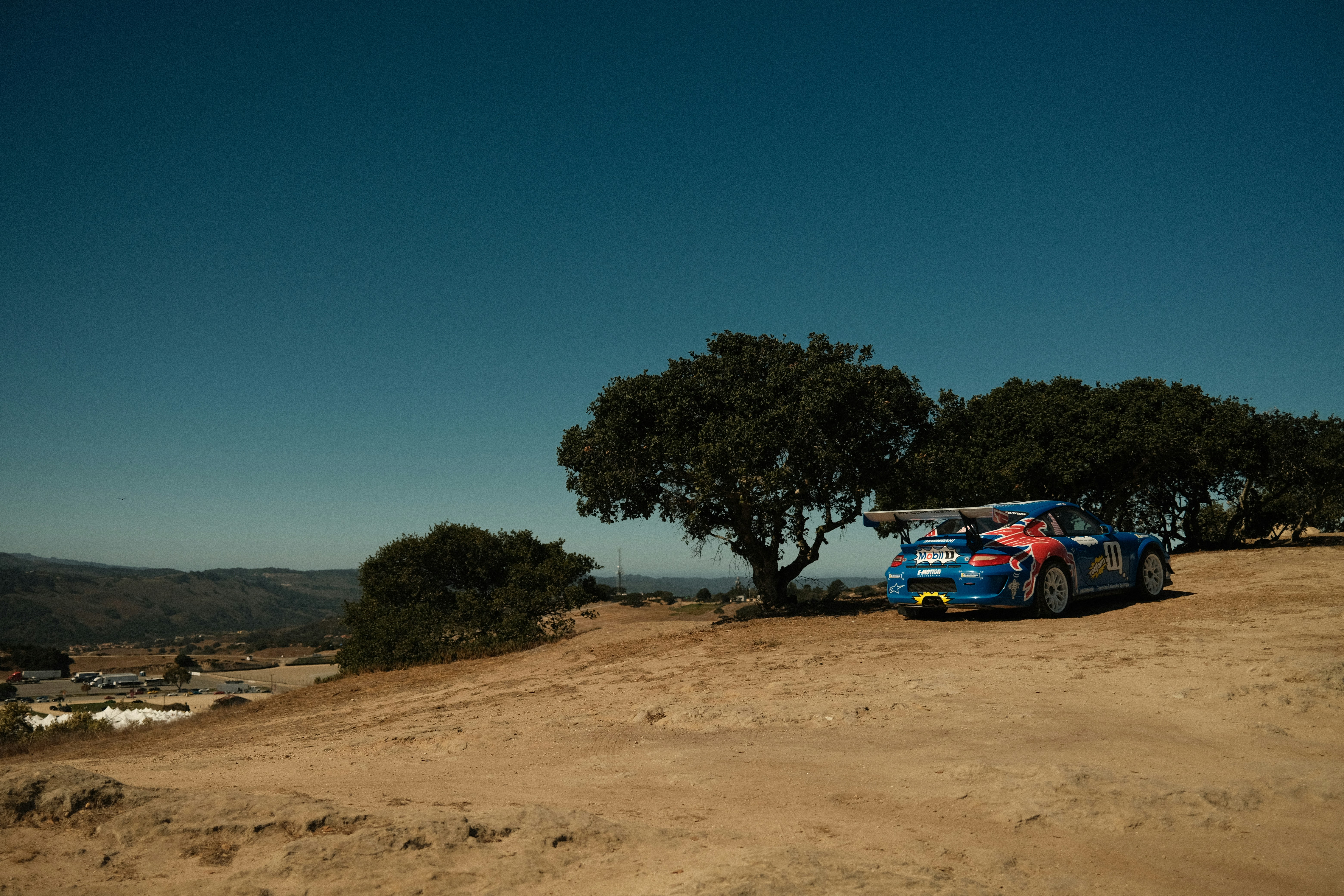 Race car parked under trees on a dusty hillside against a clear blue sky.