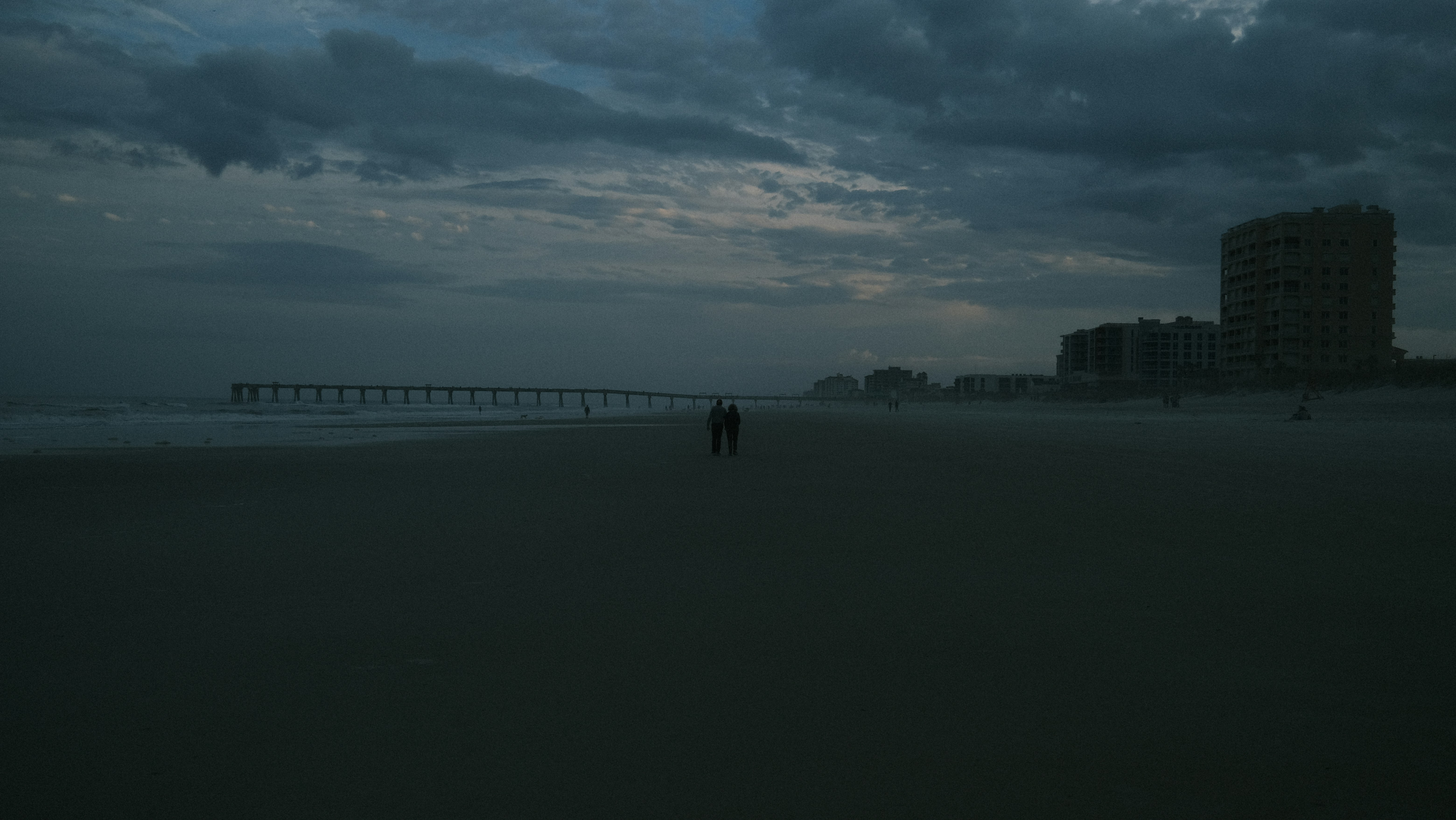 Dimly lit beach at dusk with a pier extending into the ocean and silhouettes of buildings in the distance.