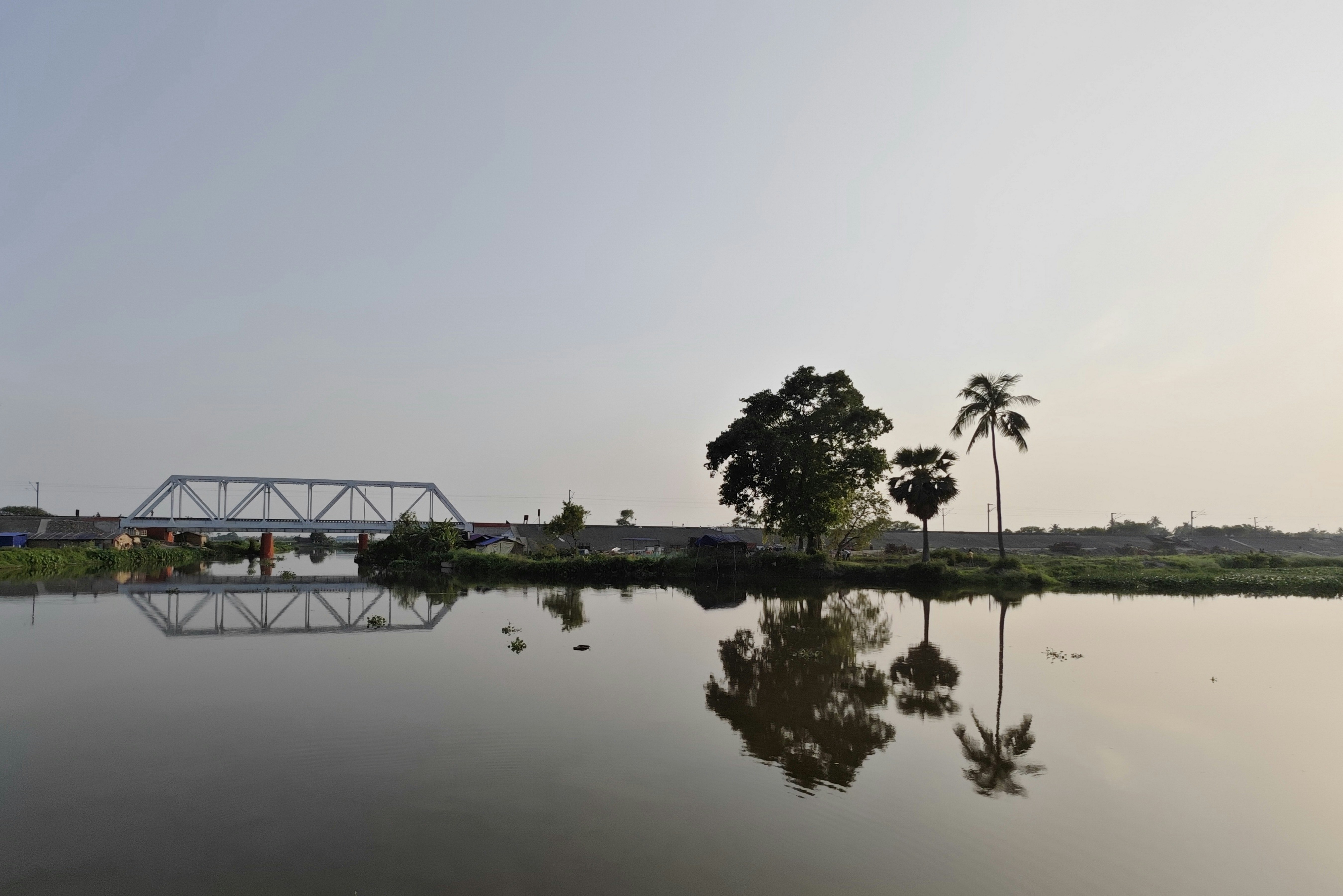 Calm riverscape photograph with a steel bridge on the left and palm trees along the bank. The still water provides a pristine reflection that doubles the scene.