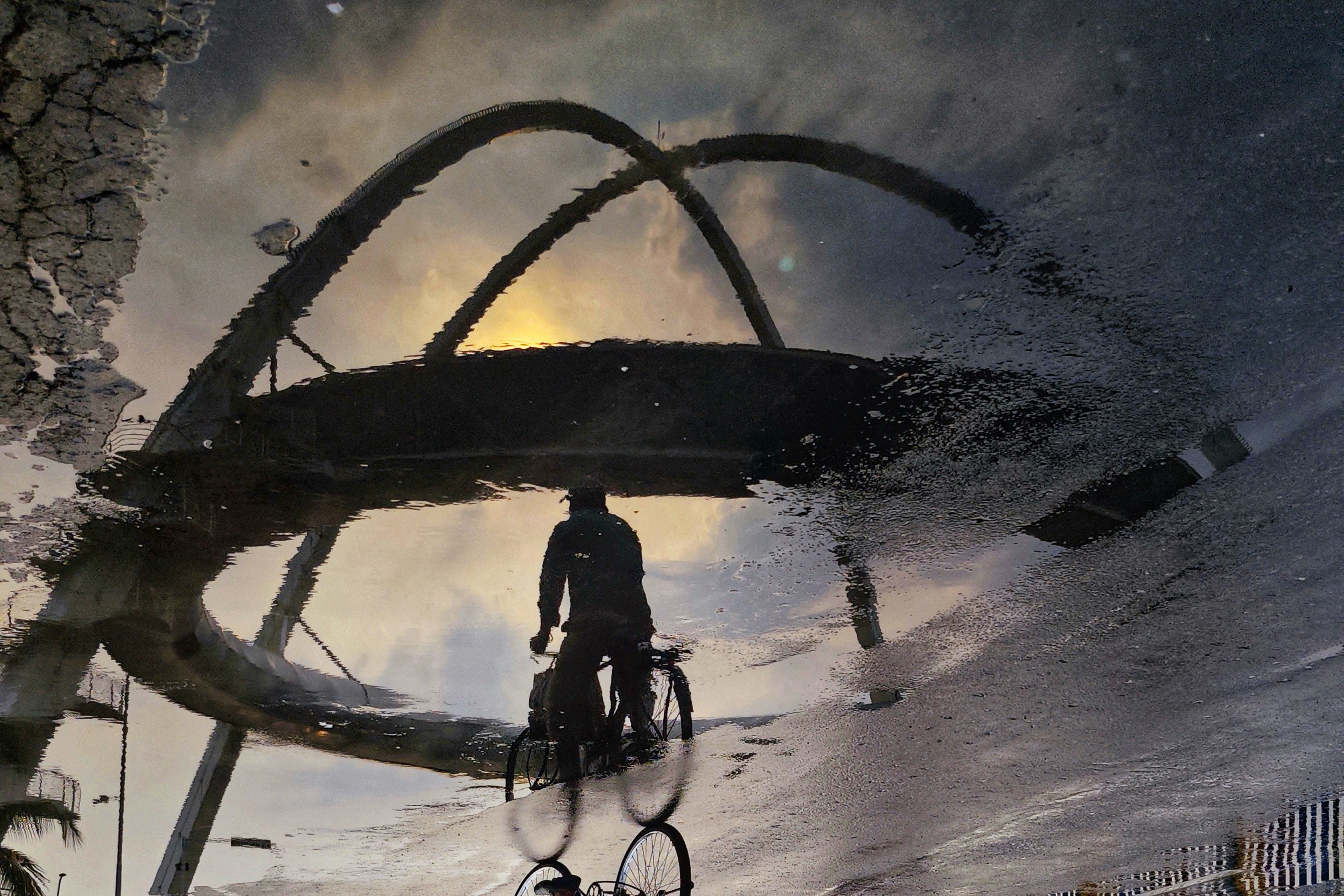 Puddle Mirror: Cyclist Framed by Looping Arches Under a Backlit Sky