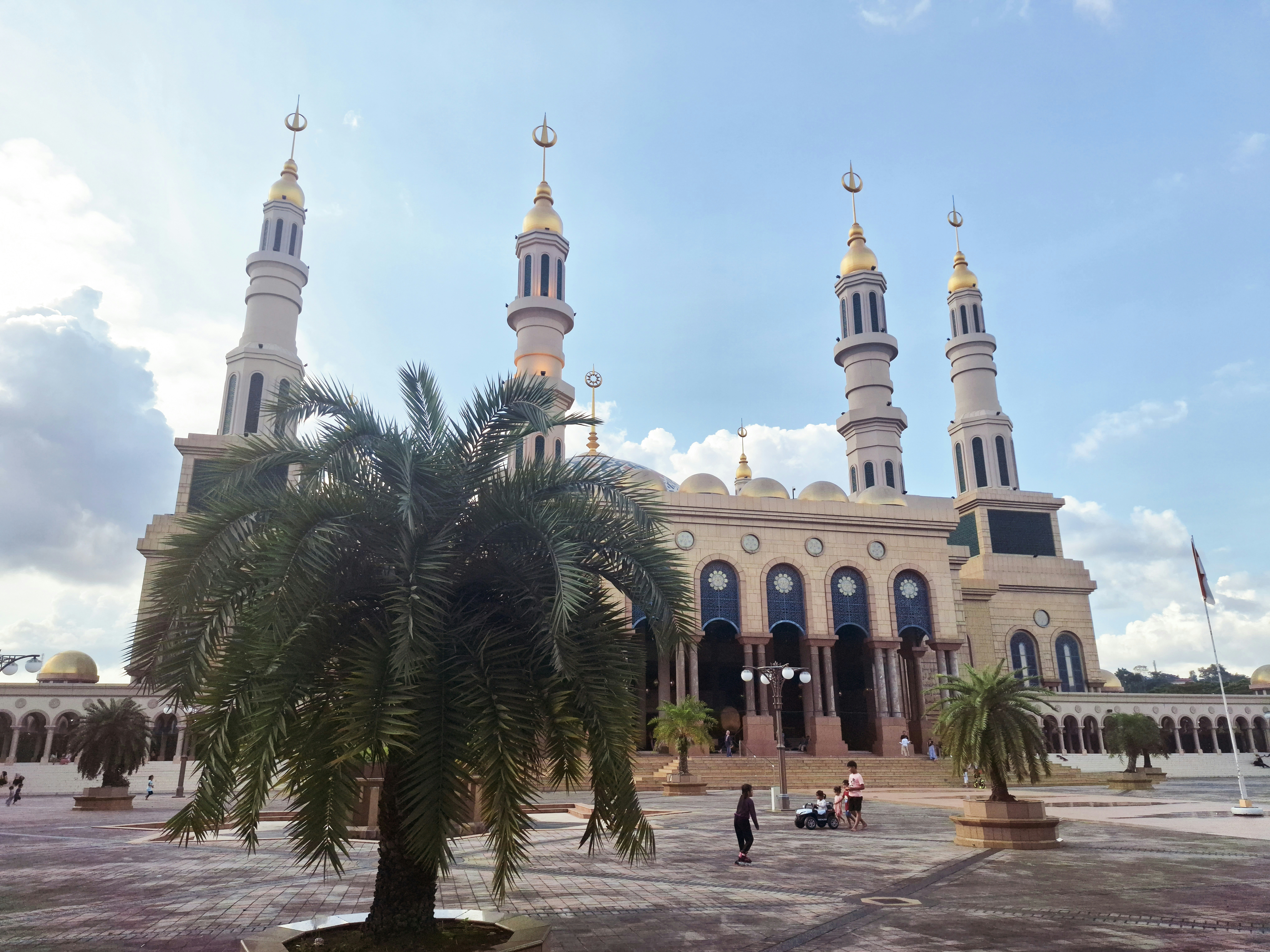 Grand mosque with four towering minarets and palm trees under a partly cloudy sky.