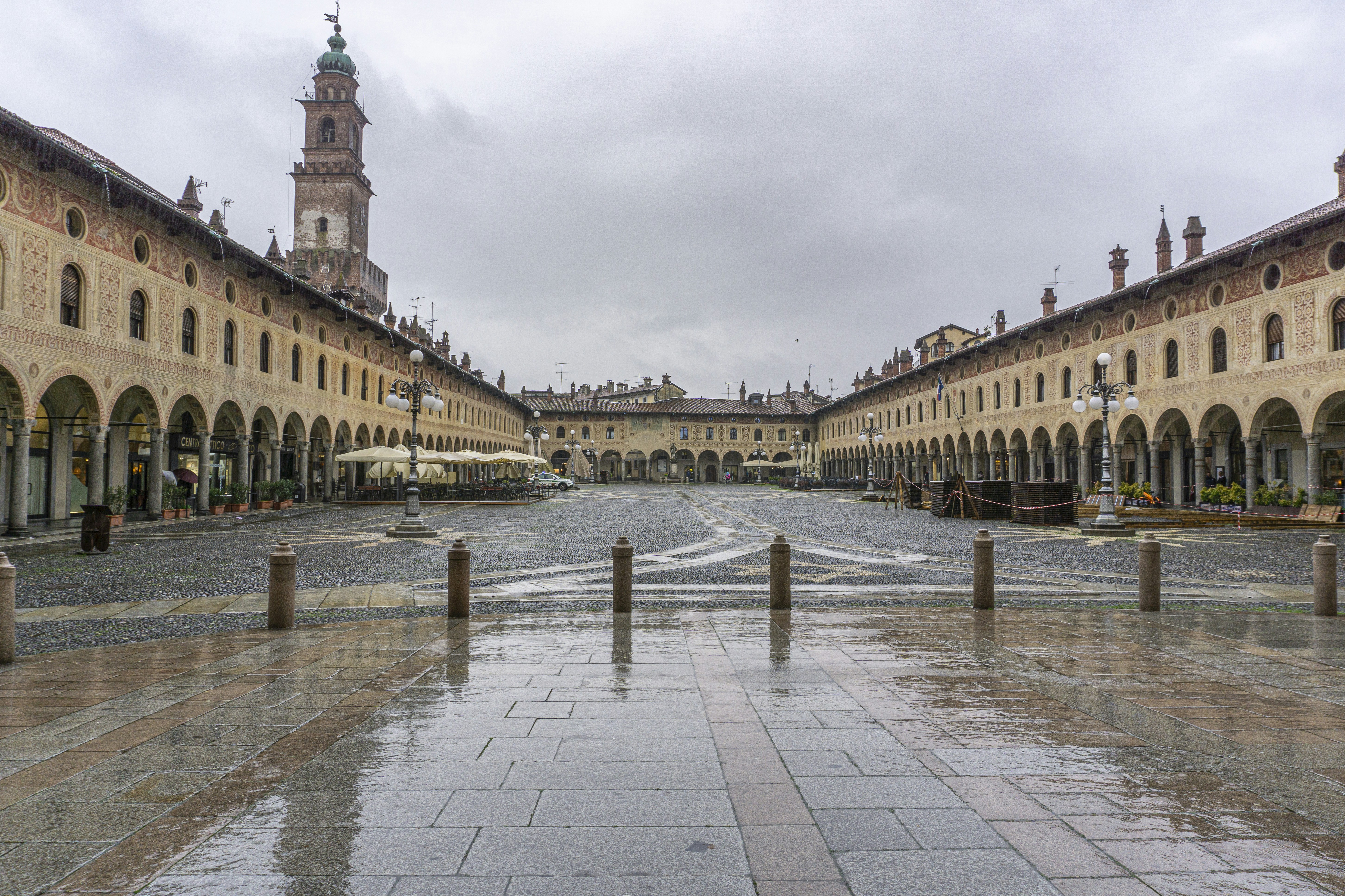 Arched colonnades line a historic piazza under an overcast sky, with reflections glistening on the wet pavement.
