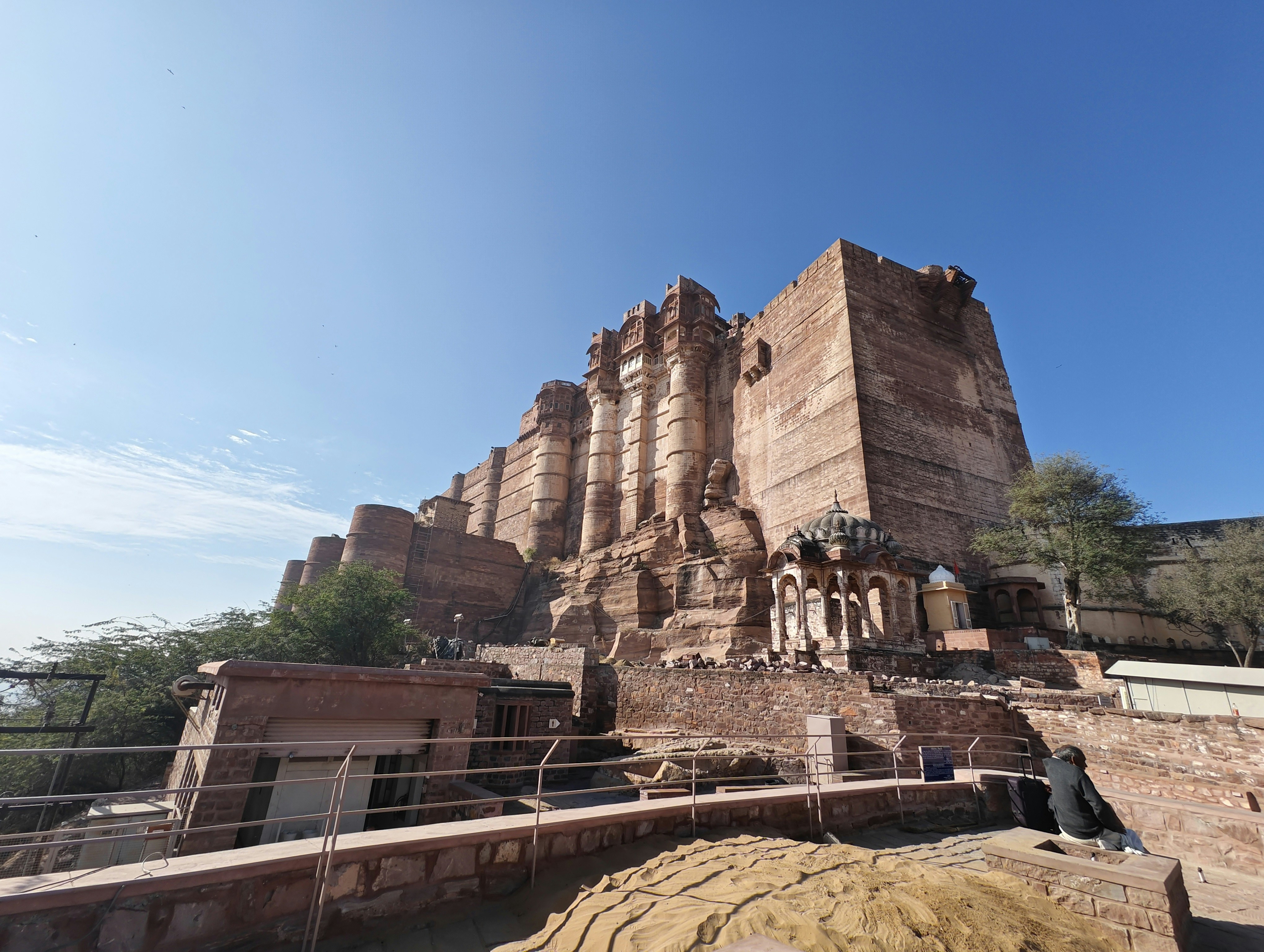 A photograph captures ancient stone fortress ruins under a clear blue sky, with a foreground viewing area and a lone visitor.