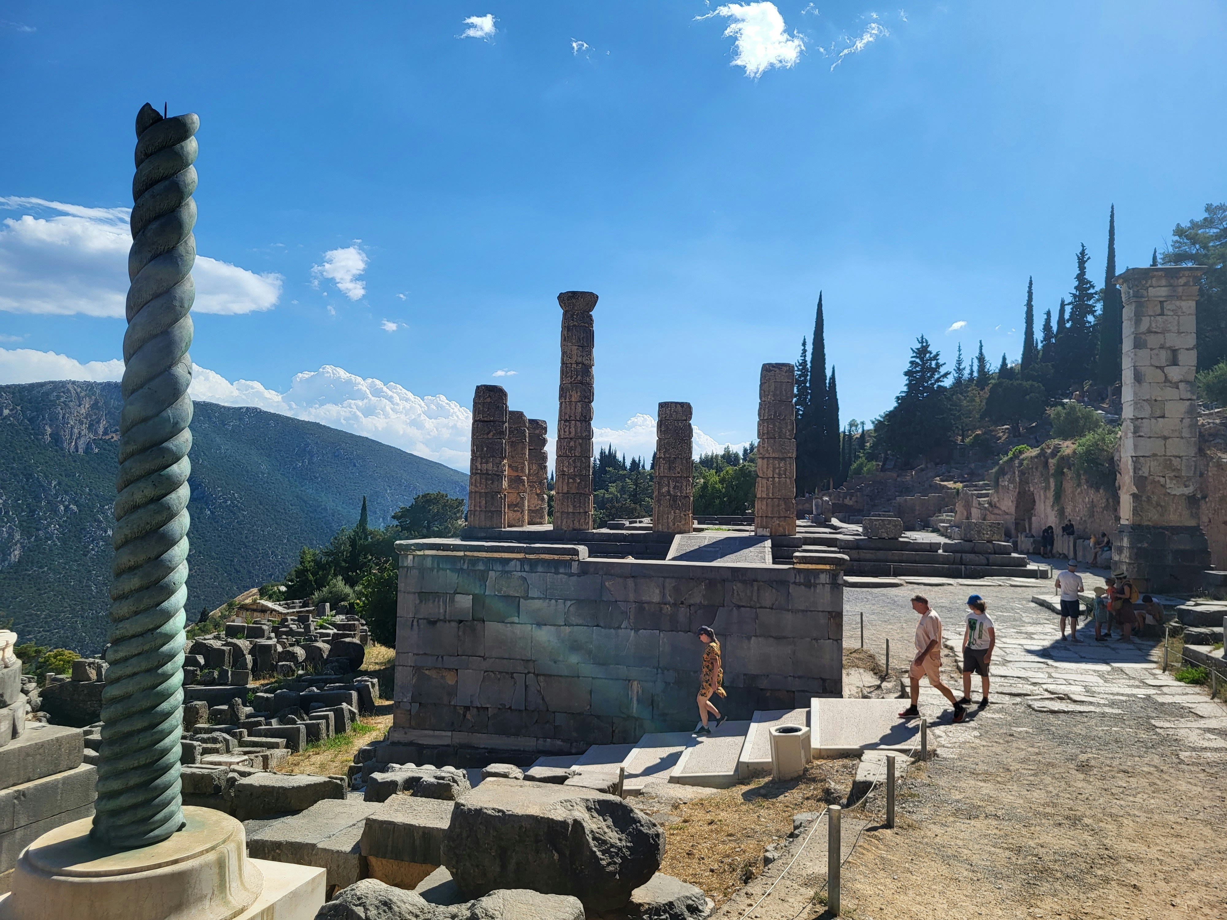 Ancient ruins of Delphi showcasing towering columns and a spiraled pillar against a backdrop of mountains and blue skies.