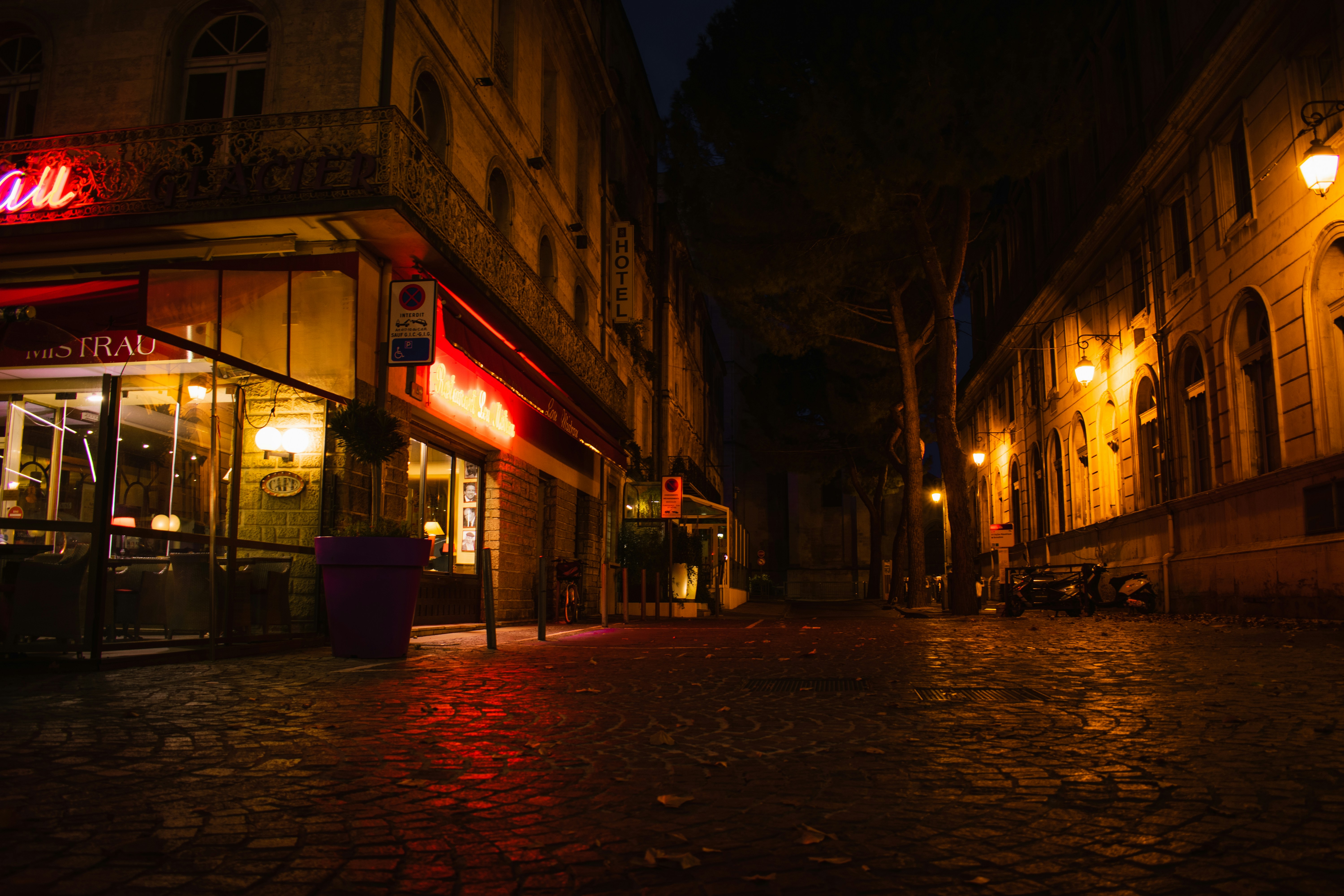 Dimly lit cobblestone street with warm glow from street lamps and café signs in a quiet urban setting.