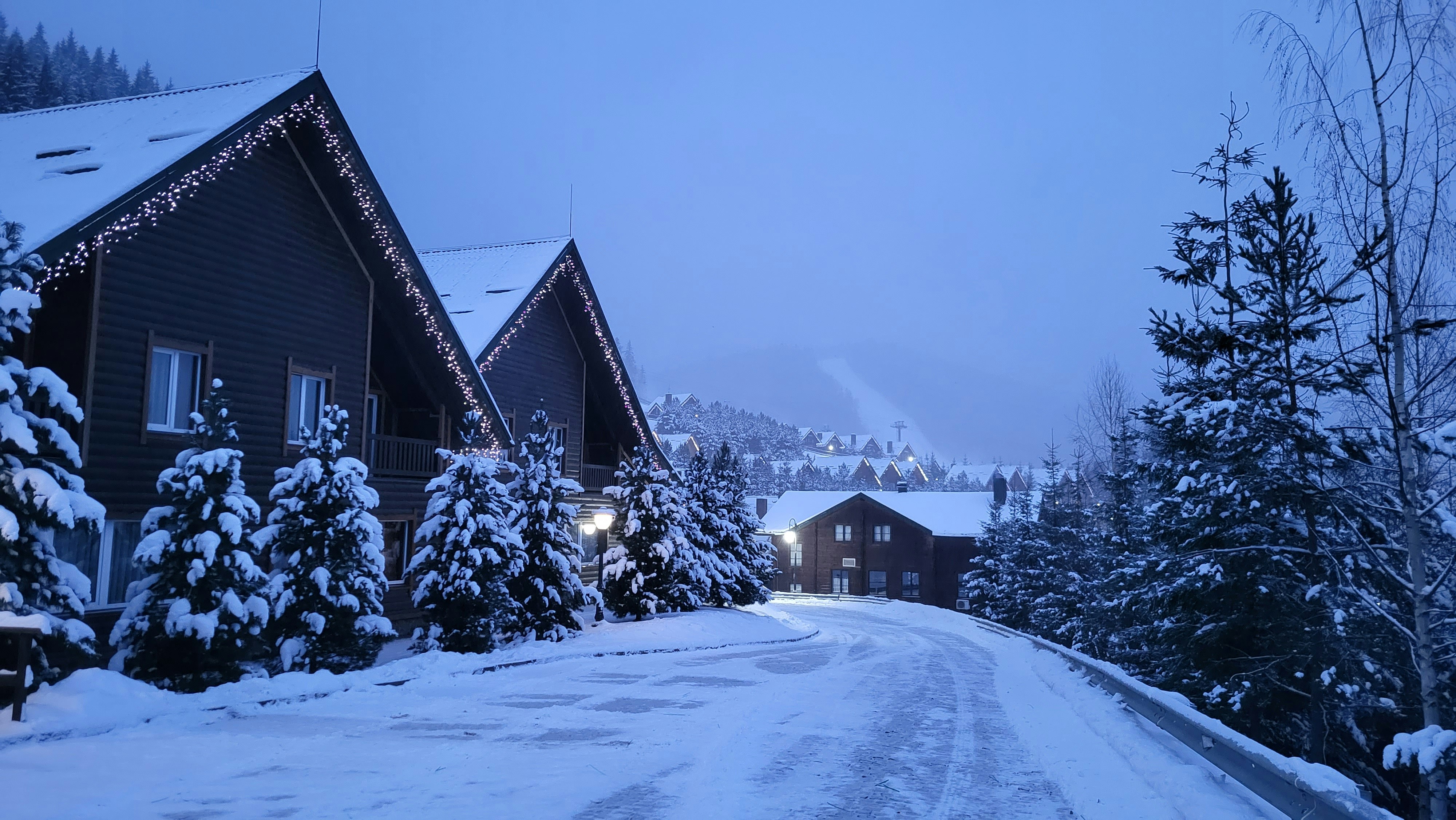 Snow-covered alpine village at blue hour with a winding road, snow-laden evergreens, and warmly lit cabins.