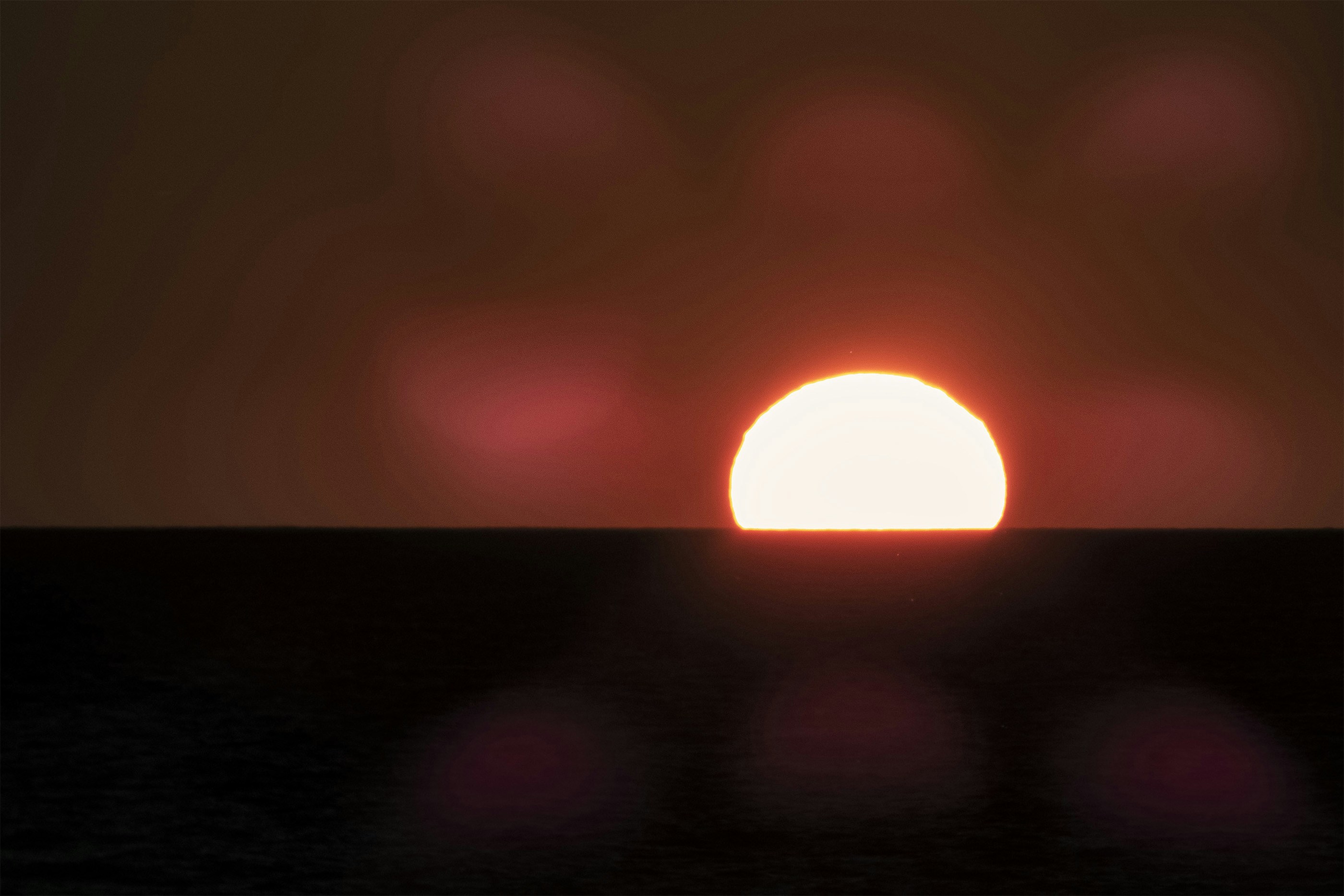 Sunset sinks behind a dark horizon over a calm sea, with large, soft bokeh circles in the foreground. This photograph captures a quiet horizon and the warm, half-lit disk of the sun.