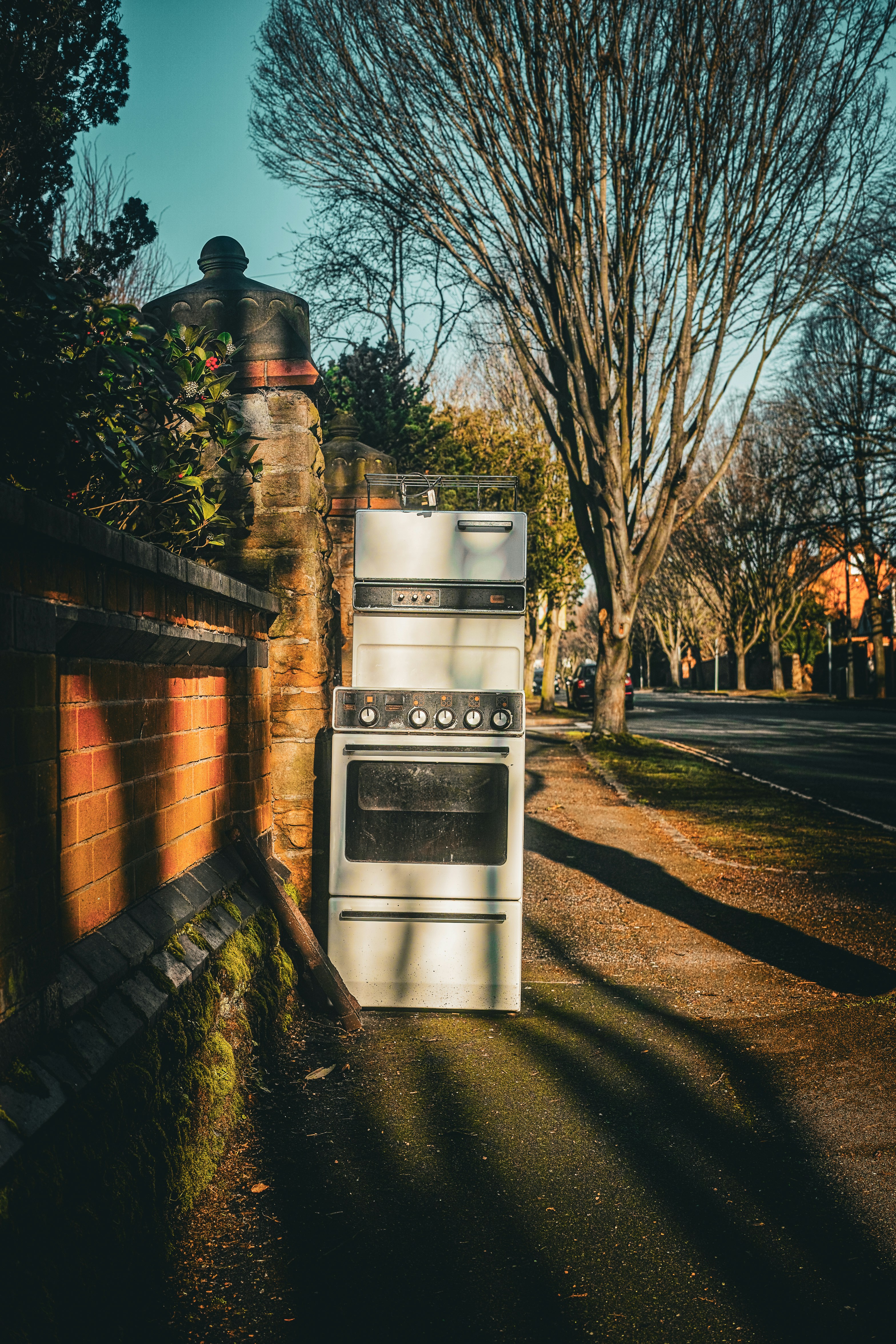A vintage freestanding oven stands upright on a sunlit sidewalk beside a brick wall, with bare trees lining a quiet suburban street.