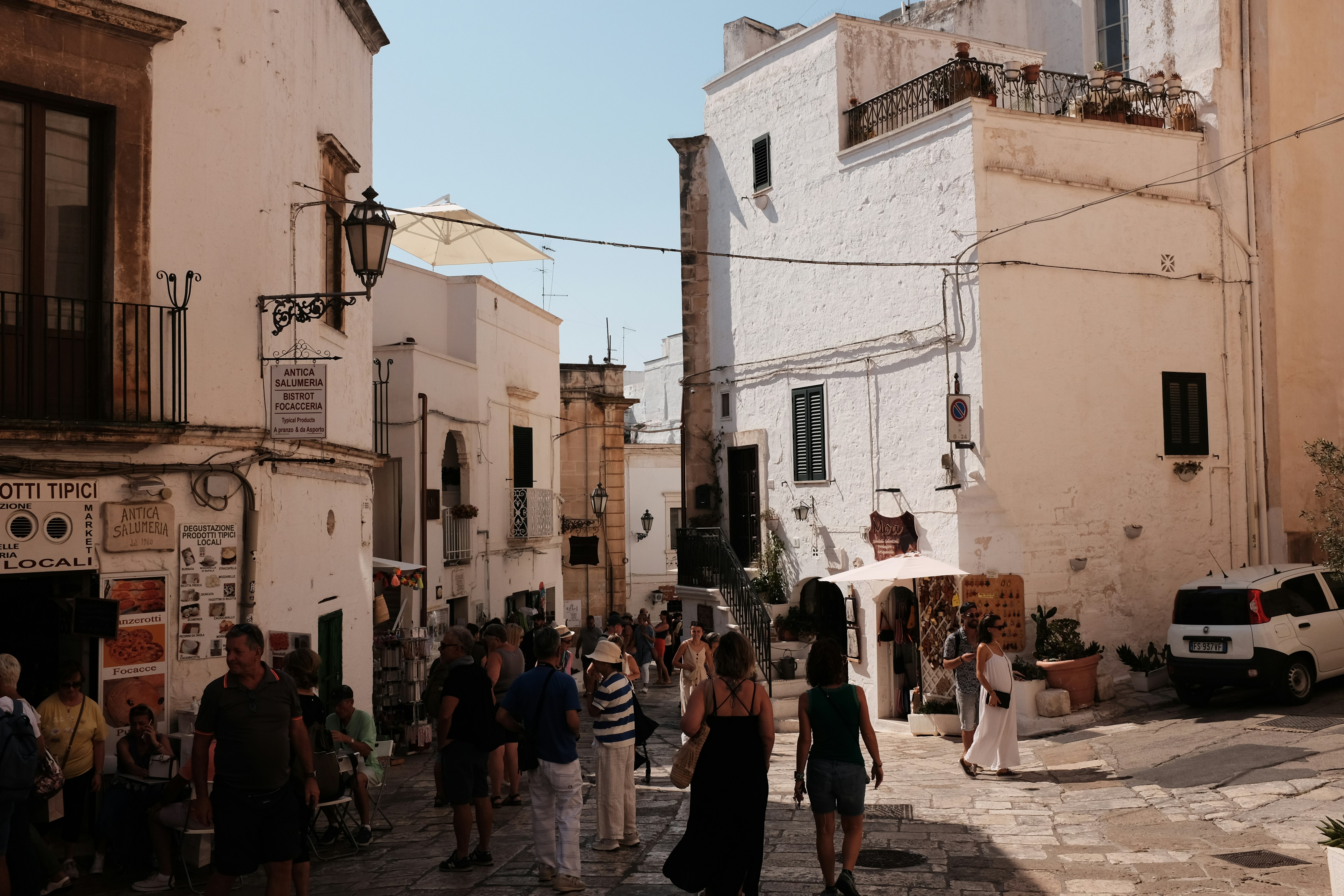 People walking along a narrow street flanked by whitewashed buildings under a clear sky.