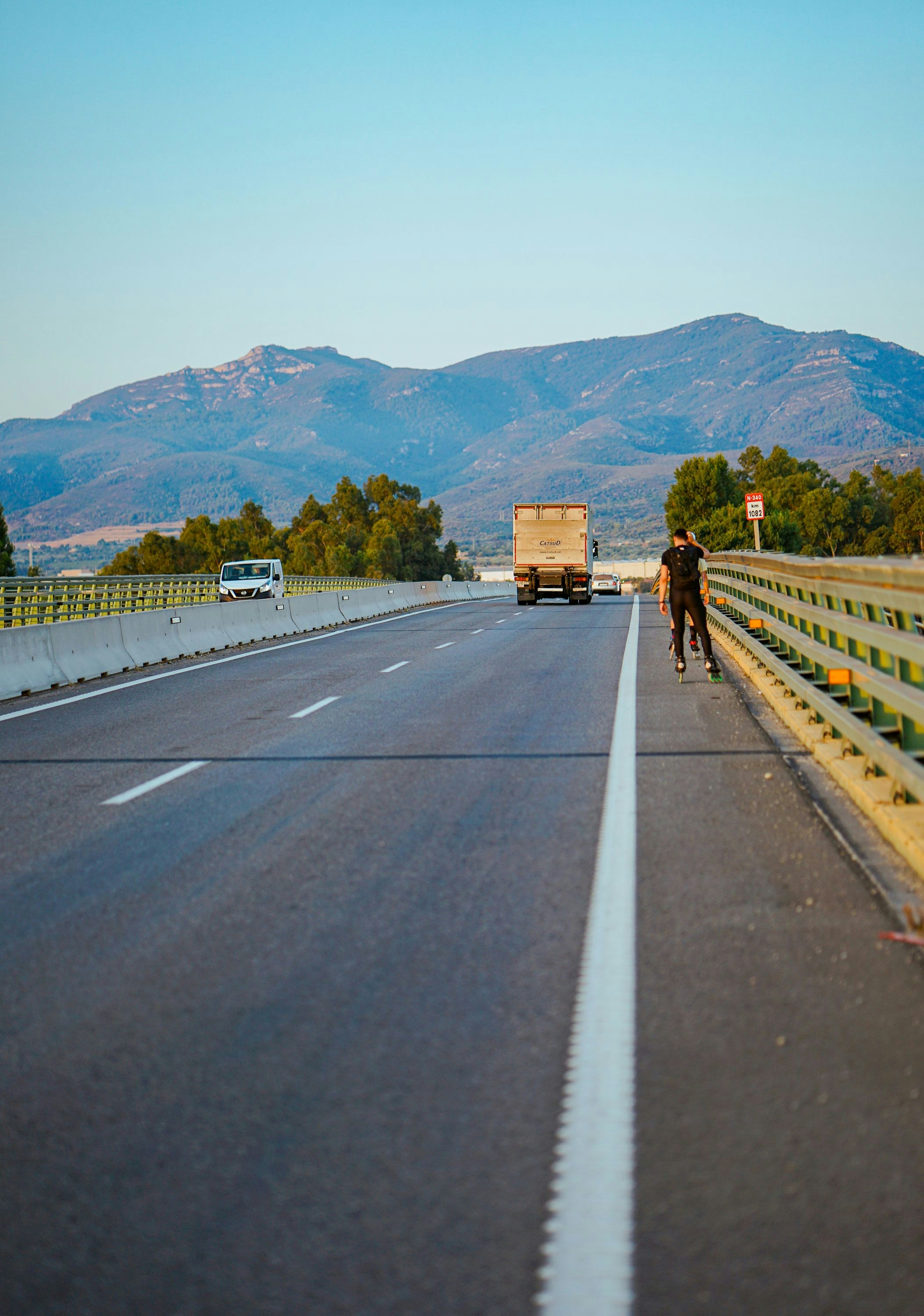 Edge of the Route: A lone cyclist on a sunlit bridge with trucks and distant peaks