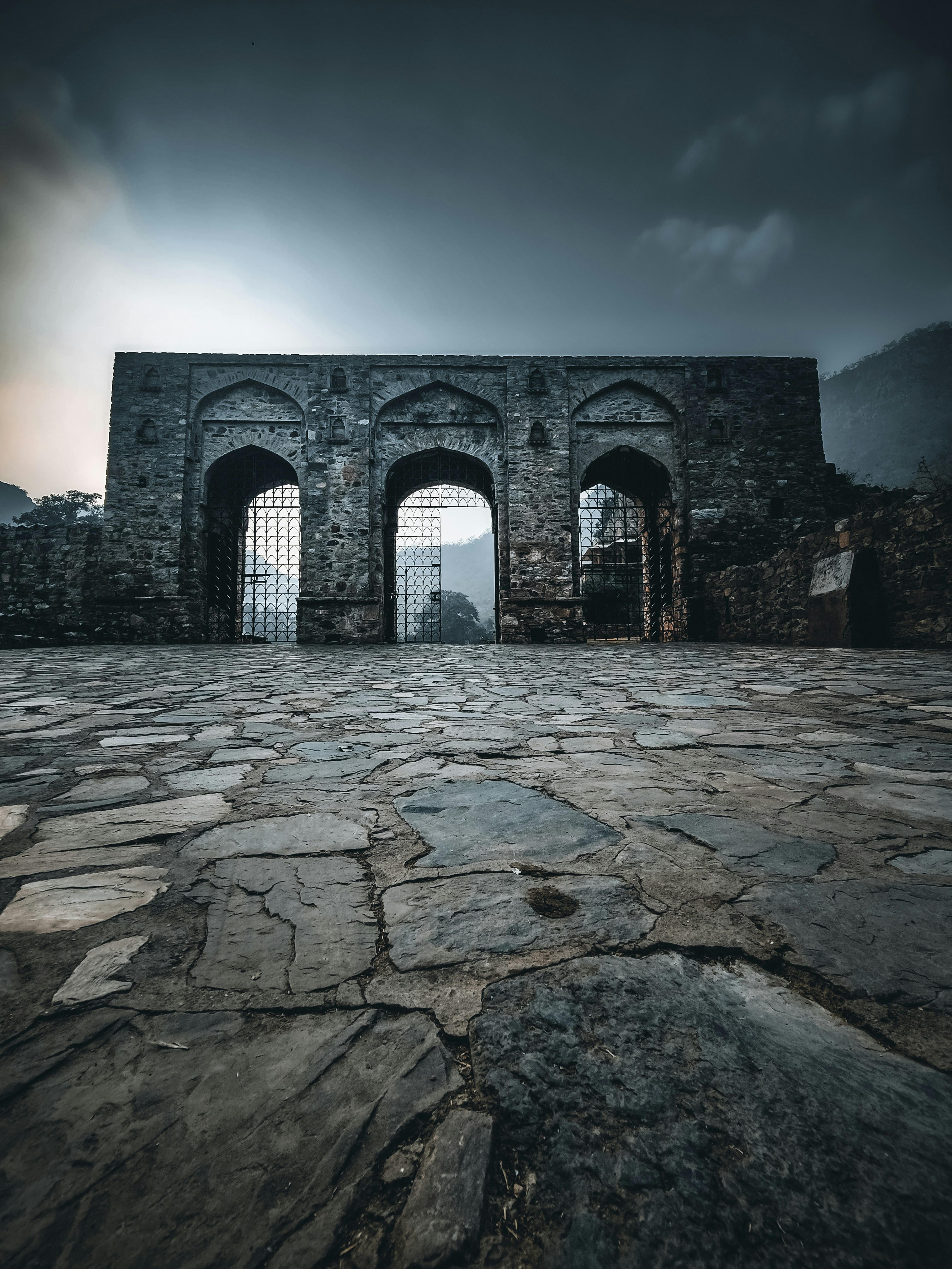 Cracked stone pavement leads to a ruined arched gateway under a moody, overcast sky.