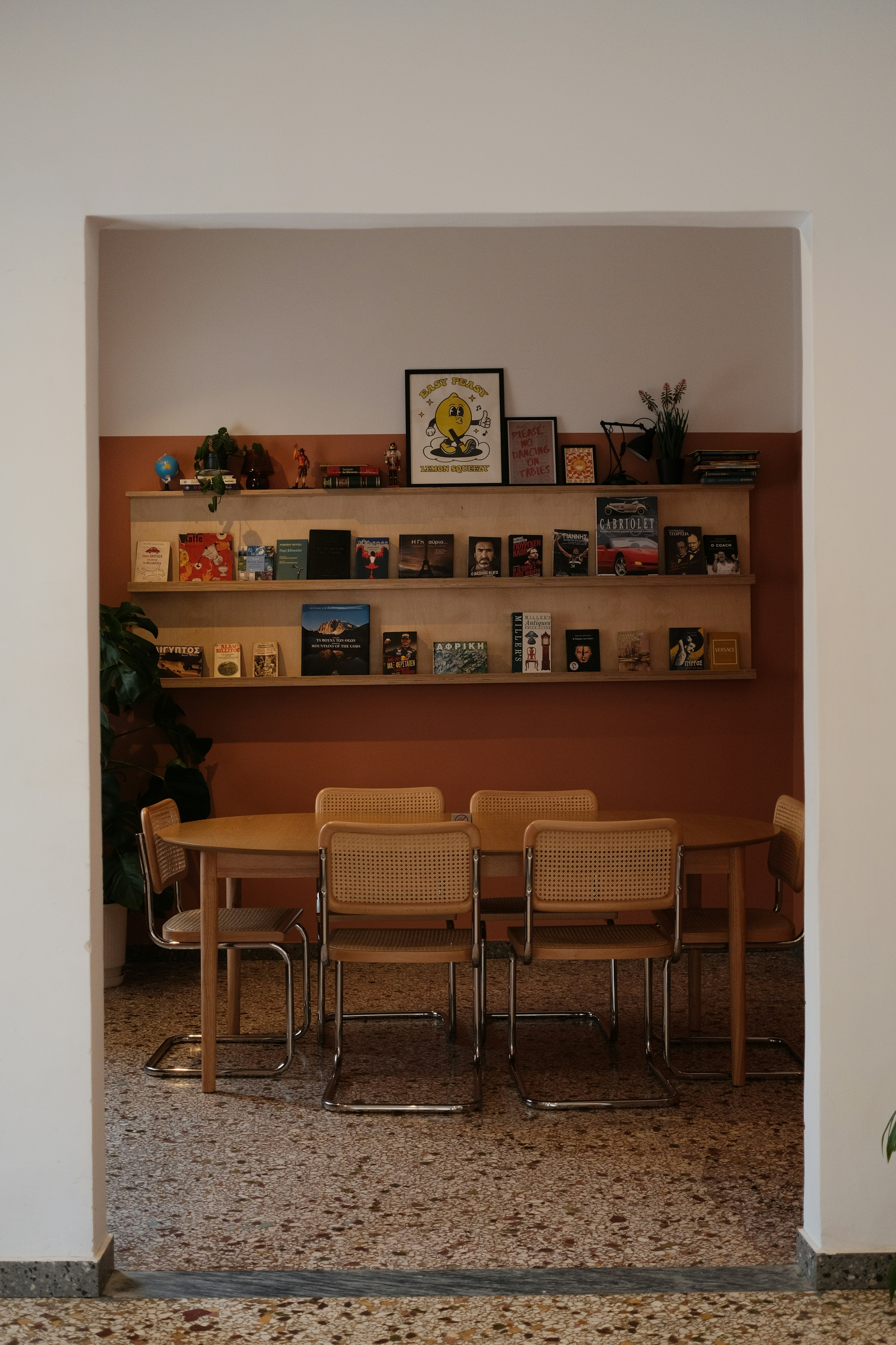 Photograph of a cozy reading nook with a round wooden table and woven chairs, set against shelves filled with books and small decor items, framed by a doorway.