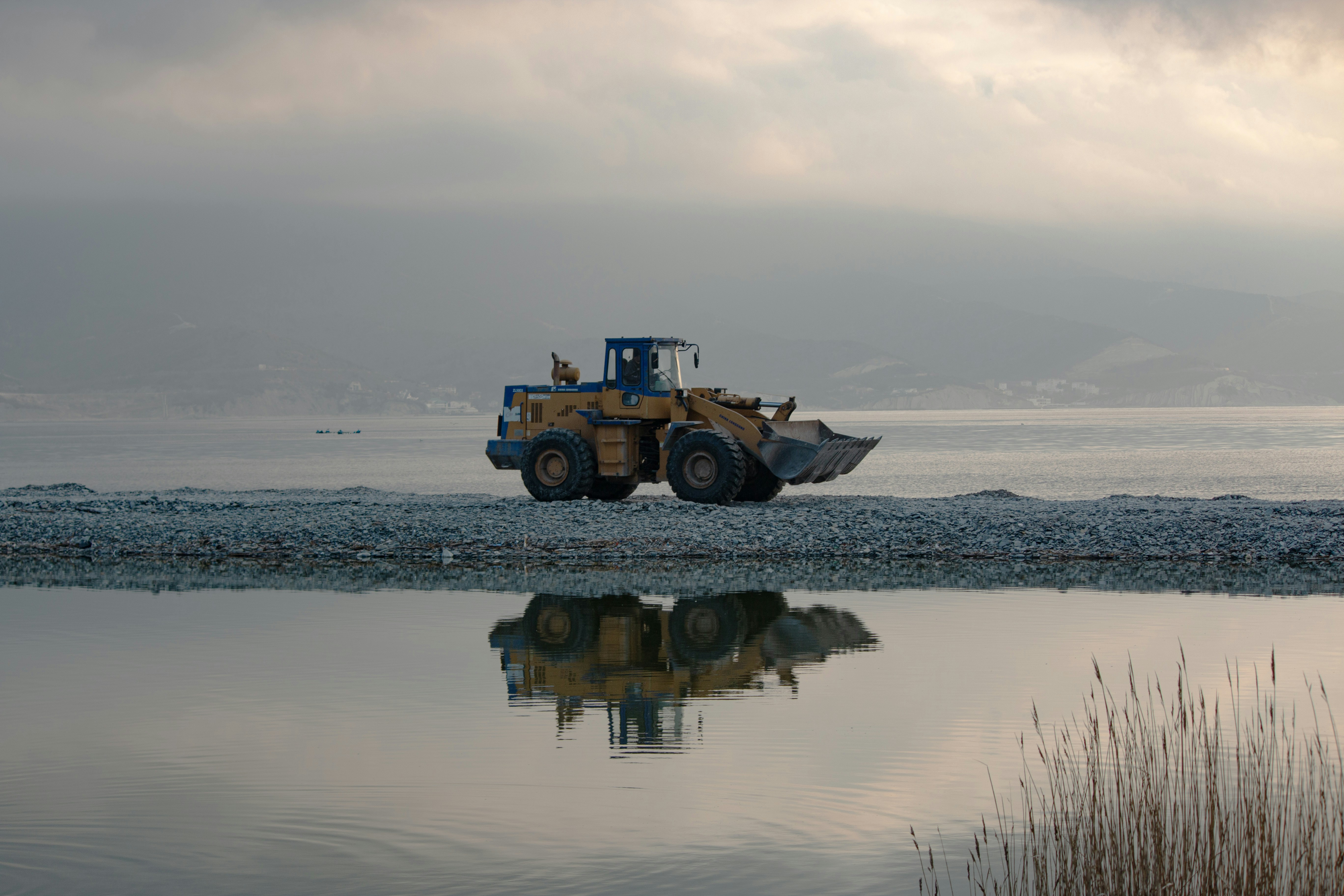 Blue excavator crossing a reflective shoreline at dusk