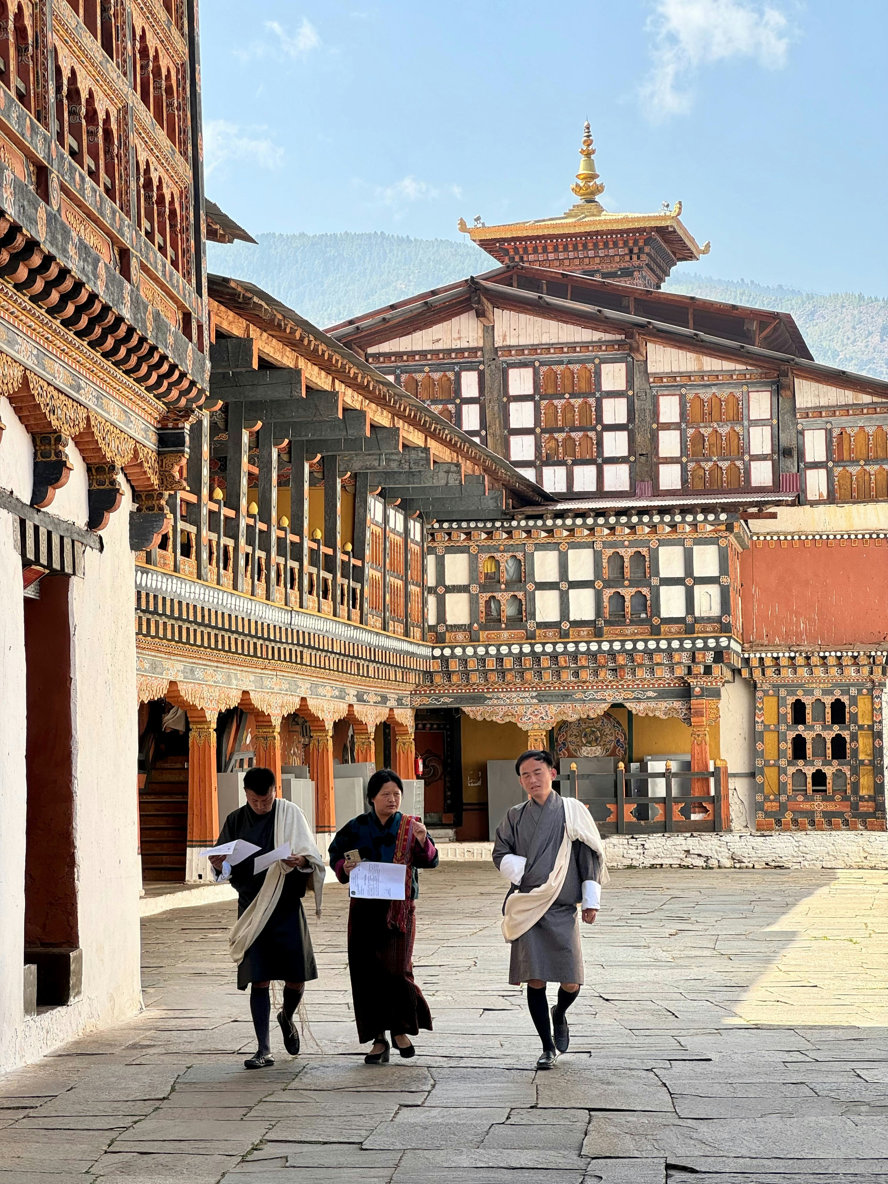 Three robed figures walk through a sunlit Himalayan monastery courtyard, framed by intricate timber-and-stucco architecture.