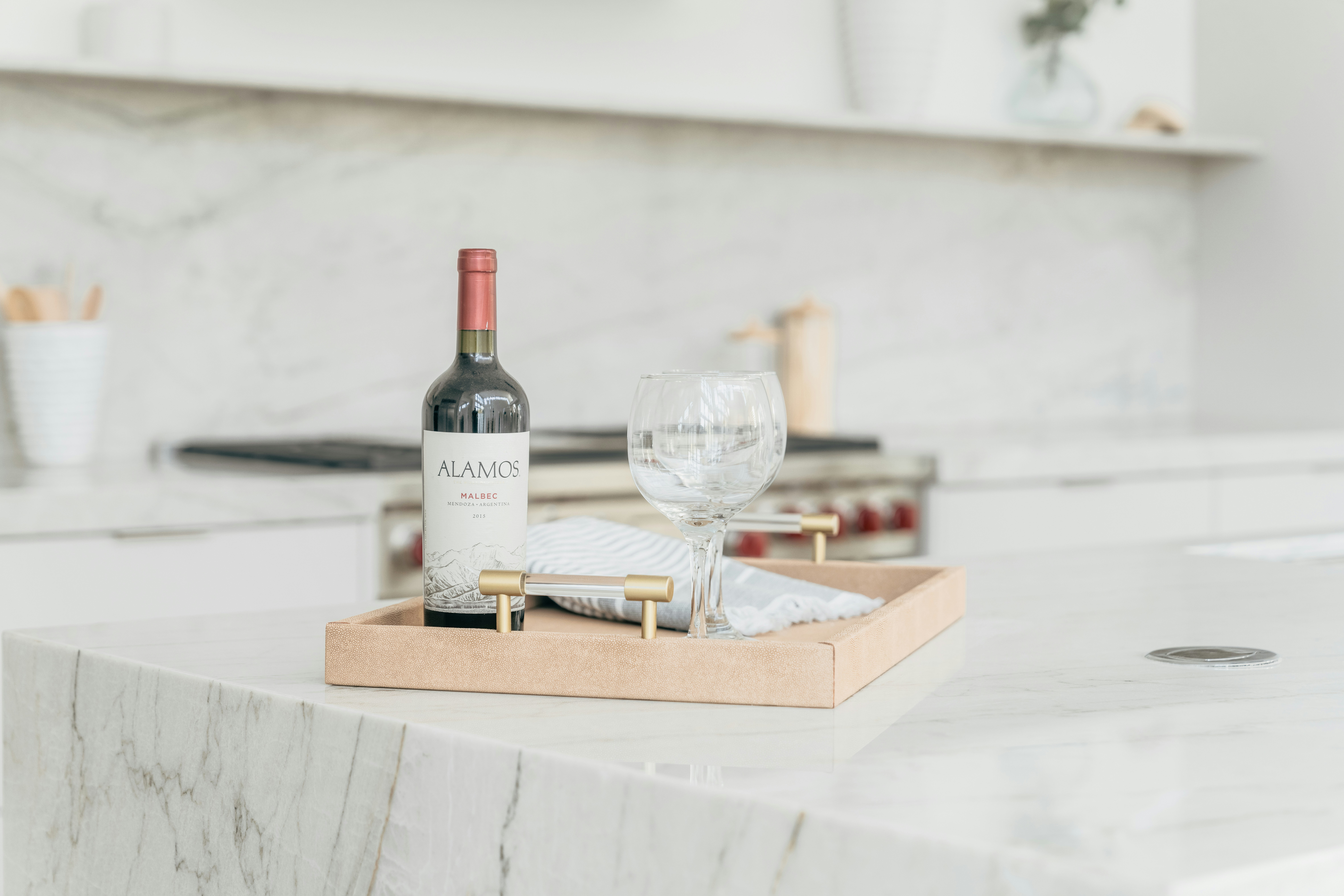 A bottle of Alamos wine and a glass rest elegantly on a wooden tray atop a marble kitchen countertop, inviting a moment of relaxation.