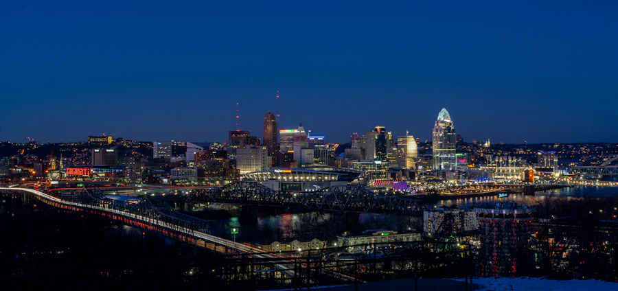 City skyline glowing at blue hour with bridges, reflections, and illuminated buildings along the river. A dark foreground frames the downtown lights.