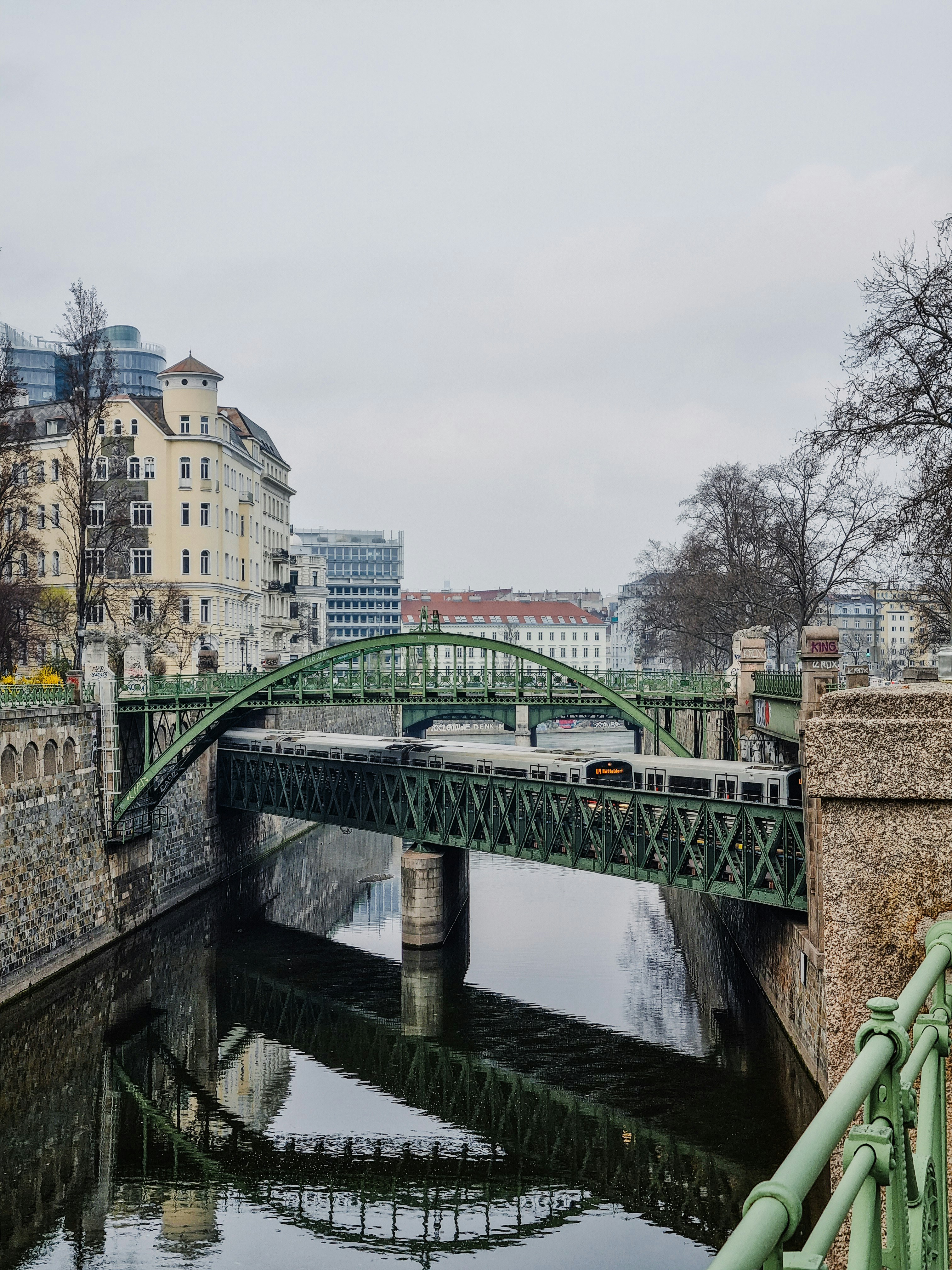 Urban river scene with a green arch bridge carrying a train, flanked by classic buildings and bare trees under a gray sky.