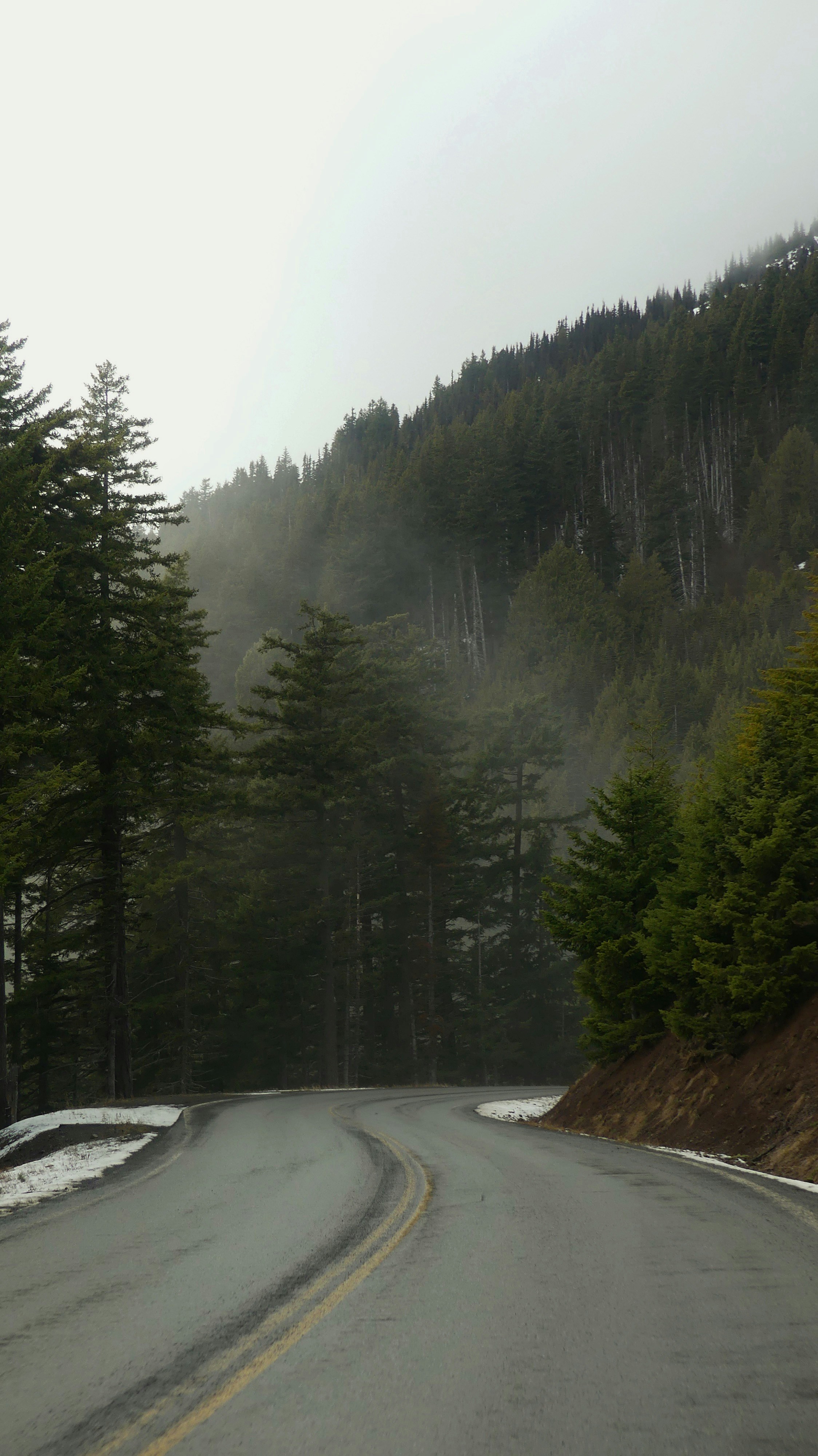 Mist-veiled Alpine Road: Snow-Lined Bend Beneath a Gentle Rainbow