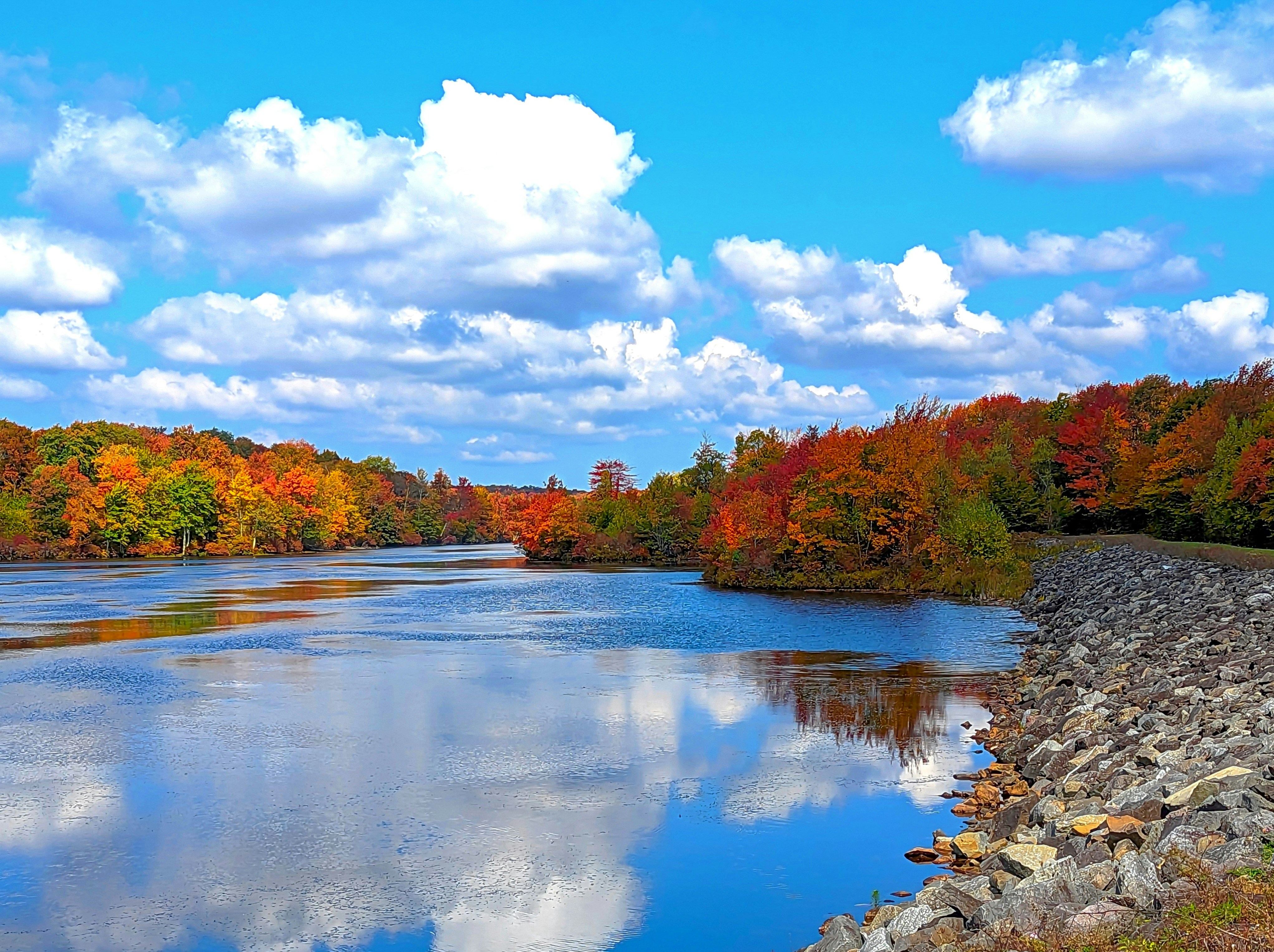 Vibrant autumn foliage mirrors in the calm waters of a river, framed by a rocky shoreline and a bright sky filled with fluffy clouds.