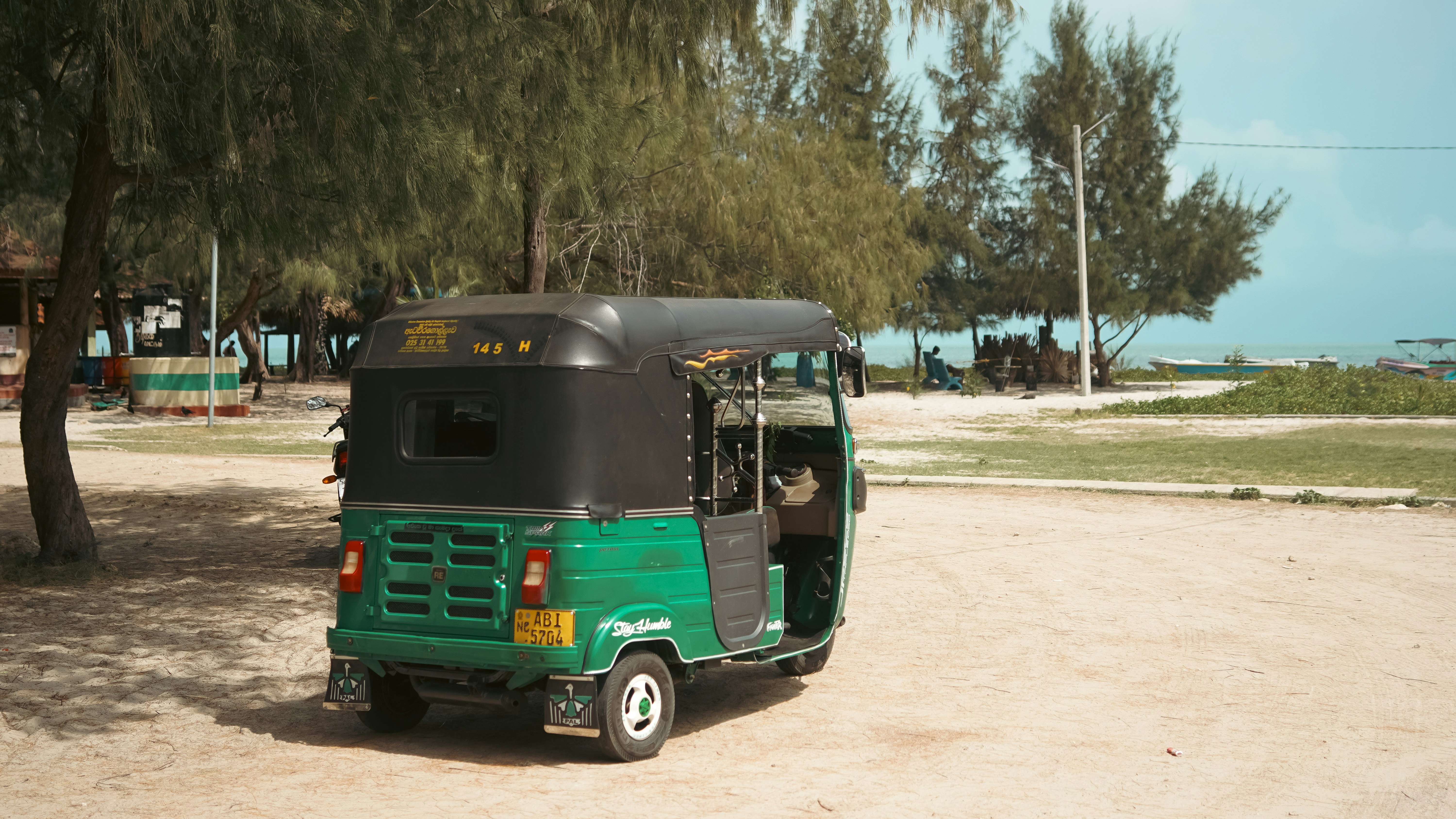 Green and black tuk-tuk parked on a sandy road near the beach with trees in the background.