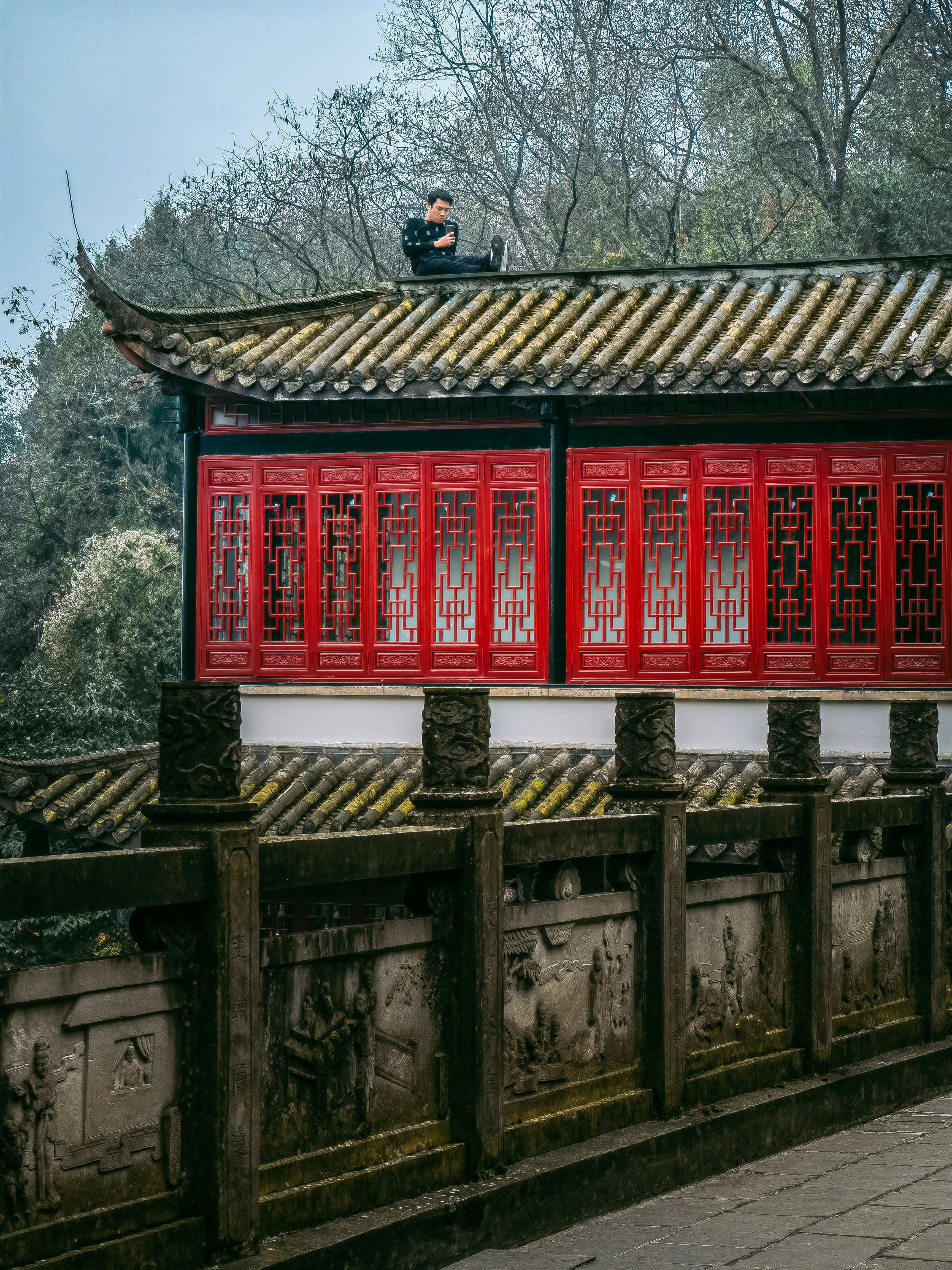 Lone figure perched on a traditional tiled roof above a crimson lattice corridor, framed by a mossy stone railing and bare trees.