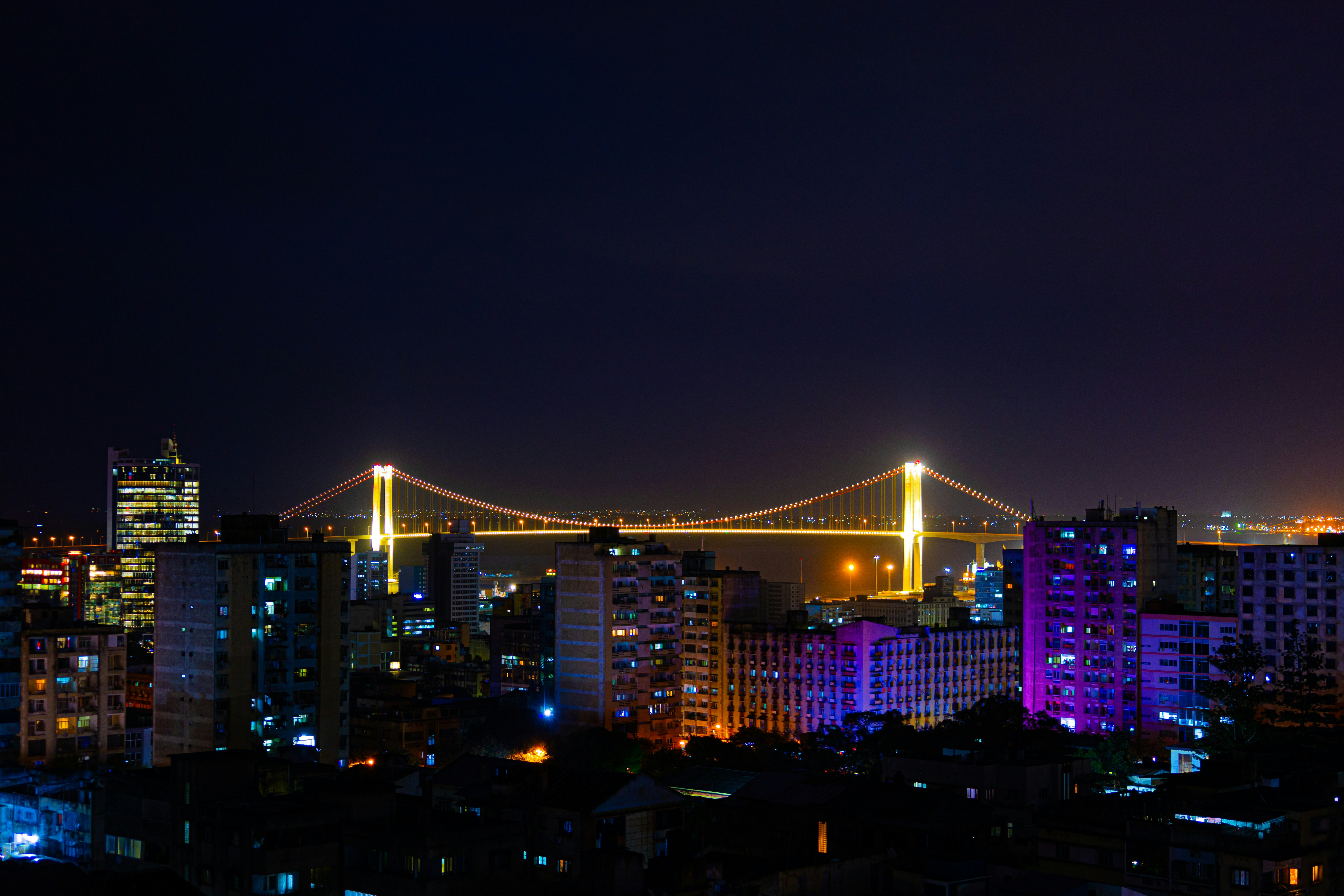 A vibrant city skyline at night featuring a beautifully lit bridge, surrounded by colorful buildings and a dark sky. The contrast highlights the urban landscape's energy.