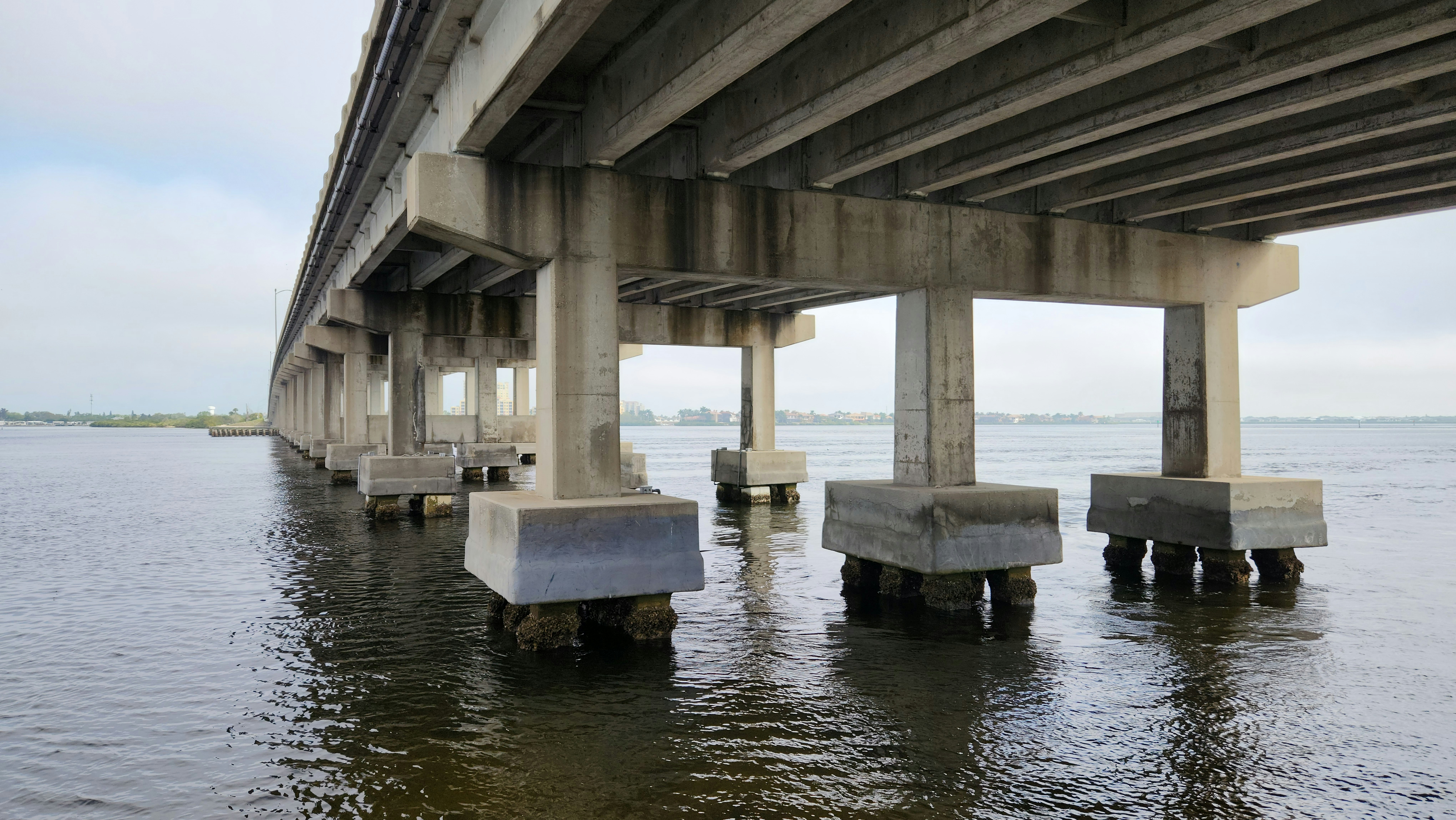 Concrete bridge piers rise from calm water beneath an elevated roadway, set against a cloudy sky. This photograph emphasizes the repetitive geometry of the columns and their reflections.
