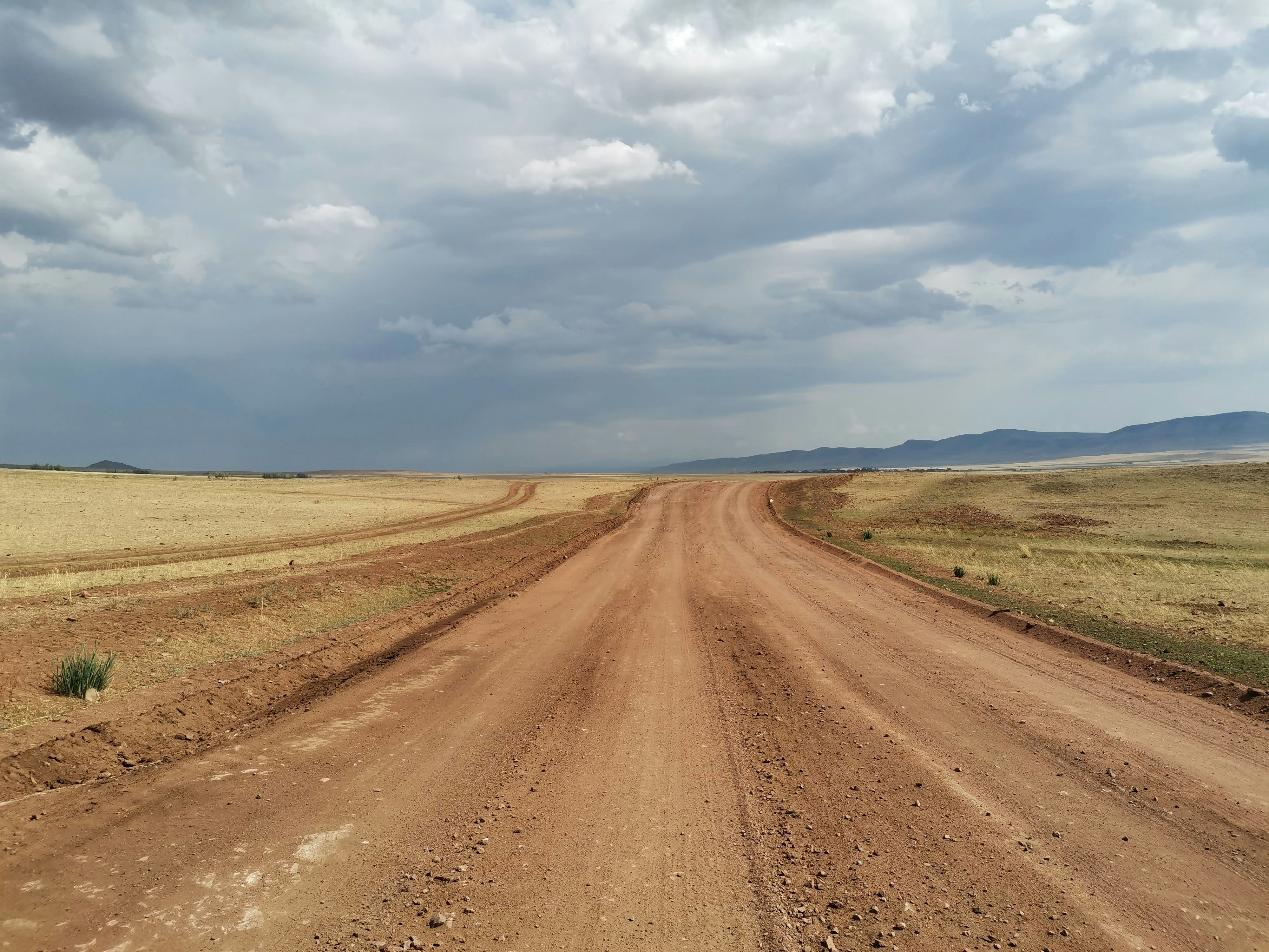 Red-dirt road stretches through open prairie toward a distant horizon under a cloudy sky.