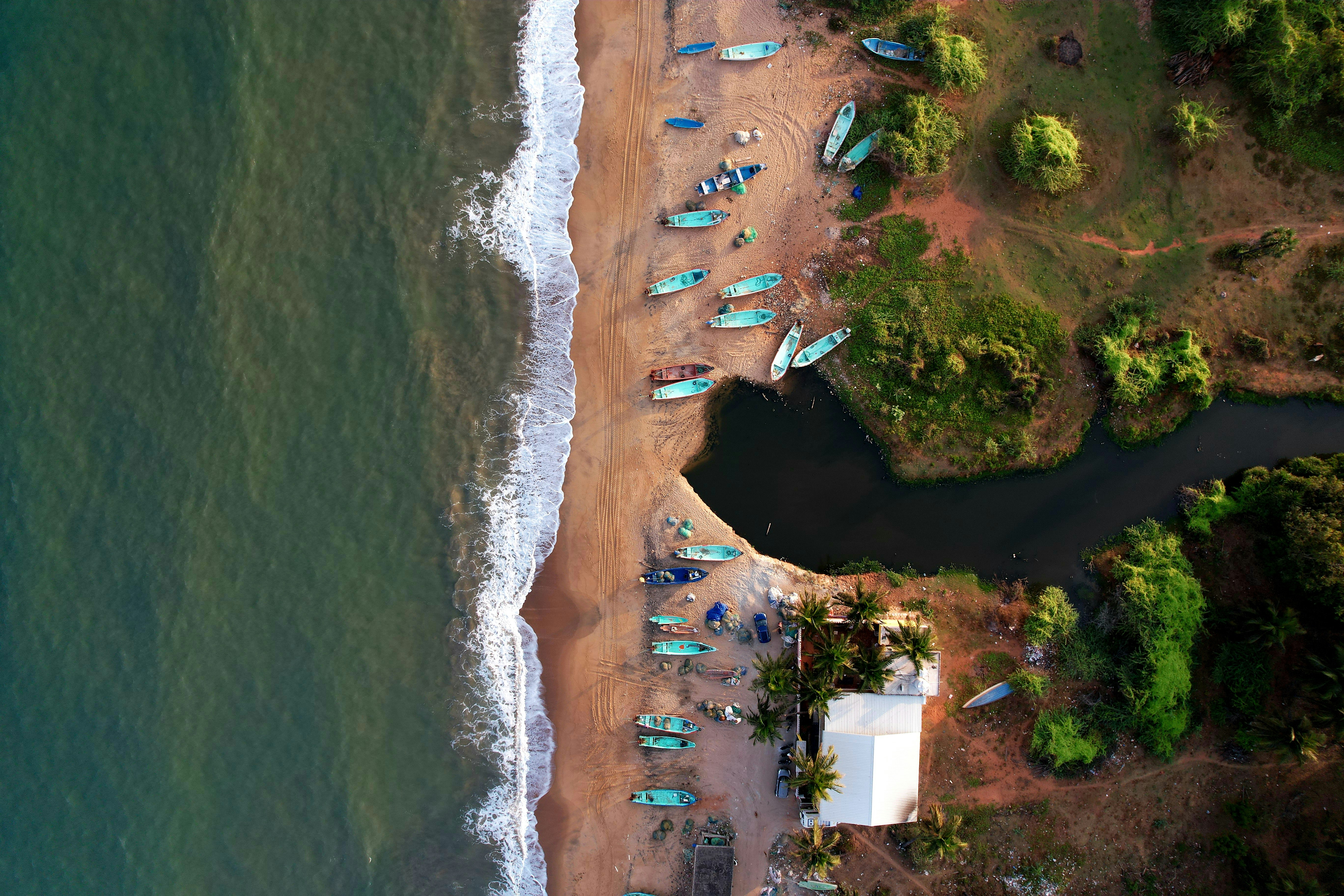 Aerial view of a sandy beach lined with colorful boats beside lush greenery and gentle ocean waves.