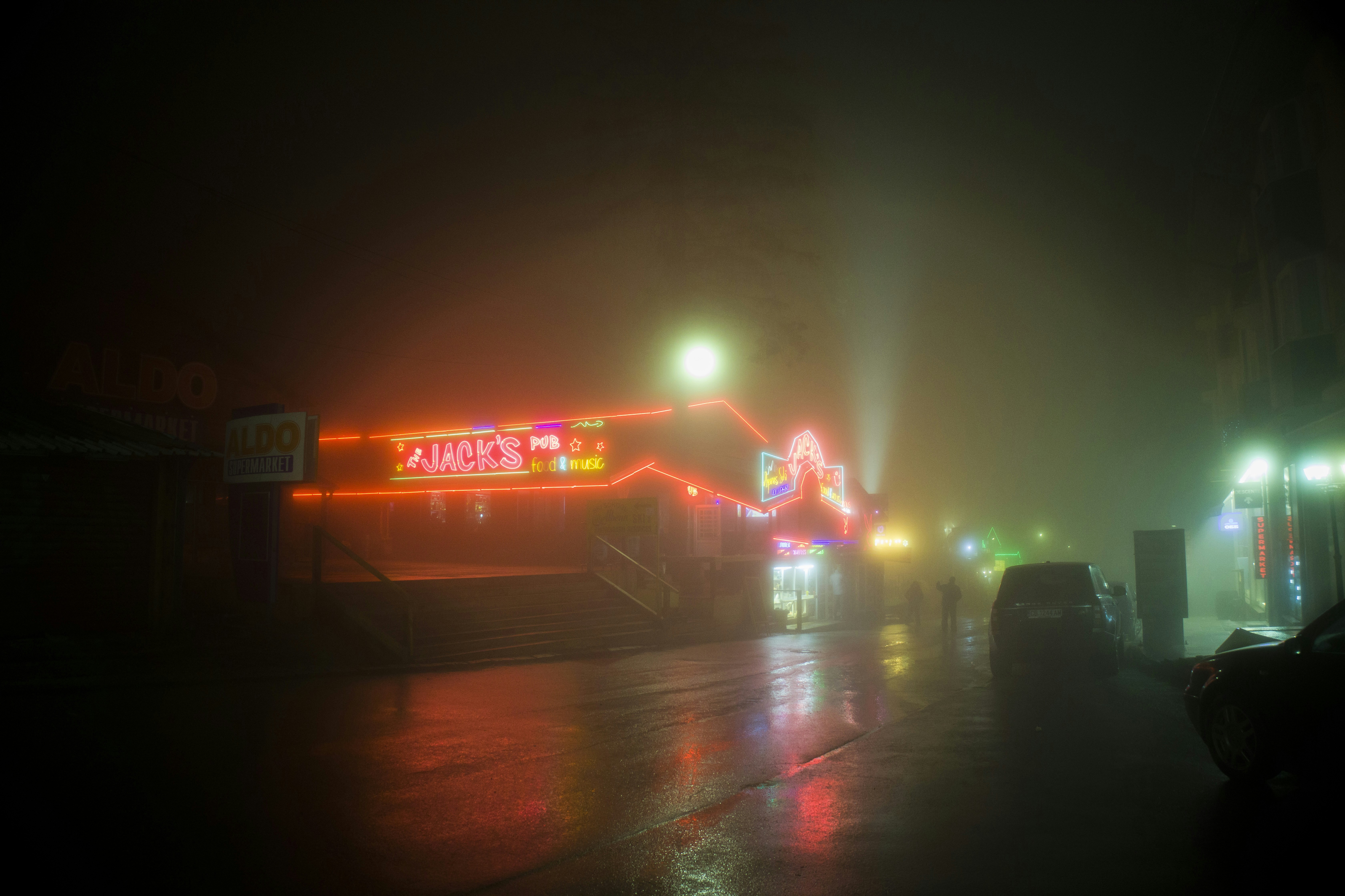 Vibrant neon signs illuminate a fog-drenched street, creating an ethereal atmosphere. The scene captures a blend of urban nightlife and atmospheric conditions.