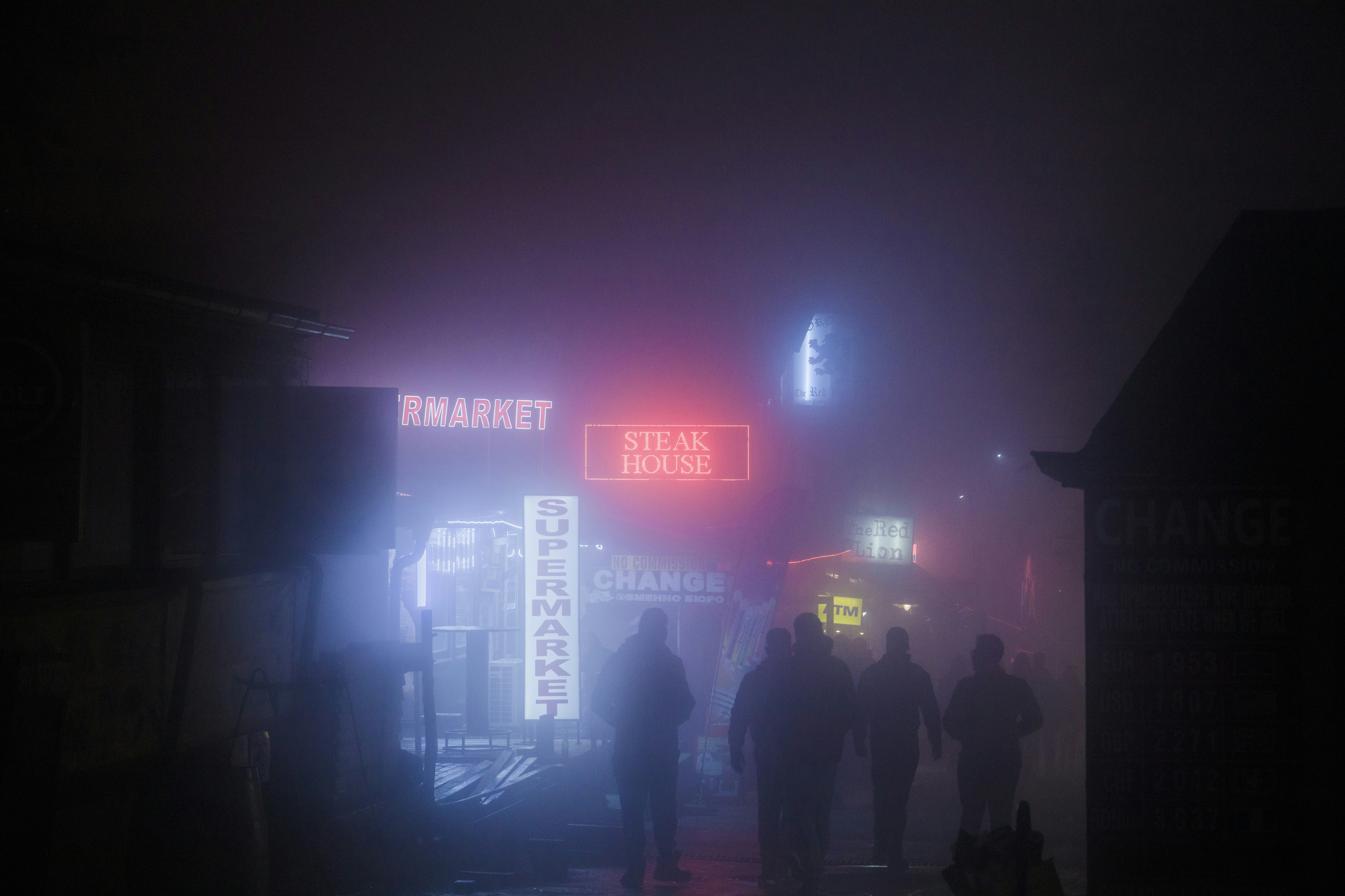 Silhouetted figures navigate a foggy street adorned with vibrant neon signs, creating an atmosphere of mystery and intrigue.
