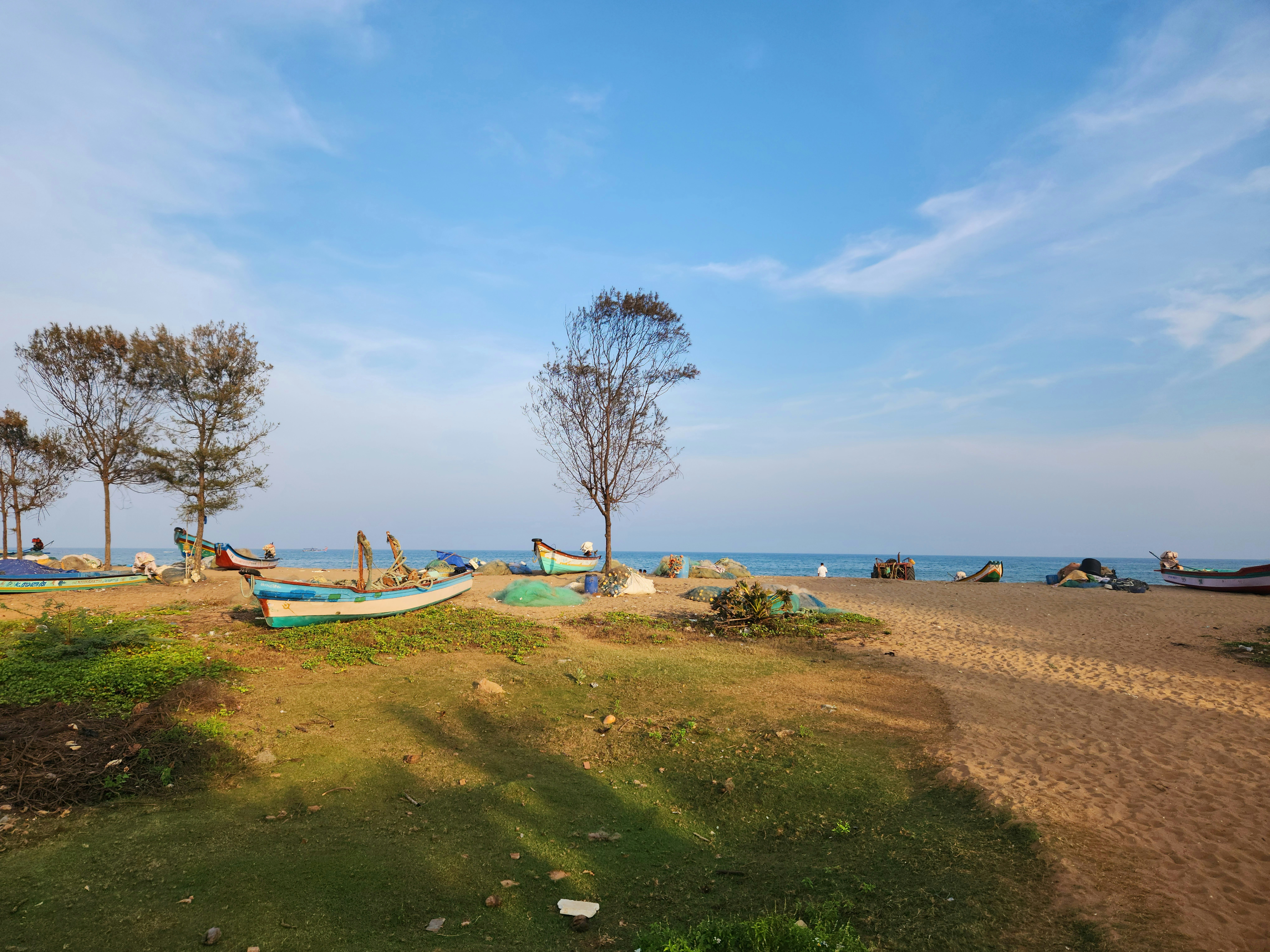 Fishing boats resting on the sandy beach under a clear blue sky, framed by sparse trees. The scene evokes a peaceful coastal atmosphere.