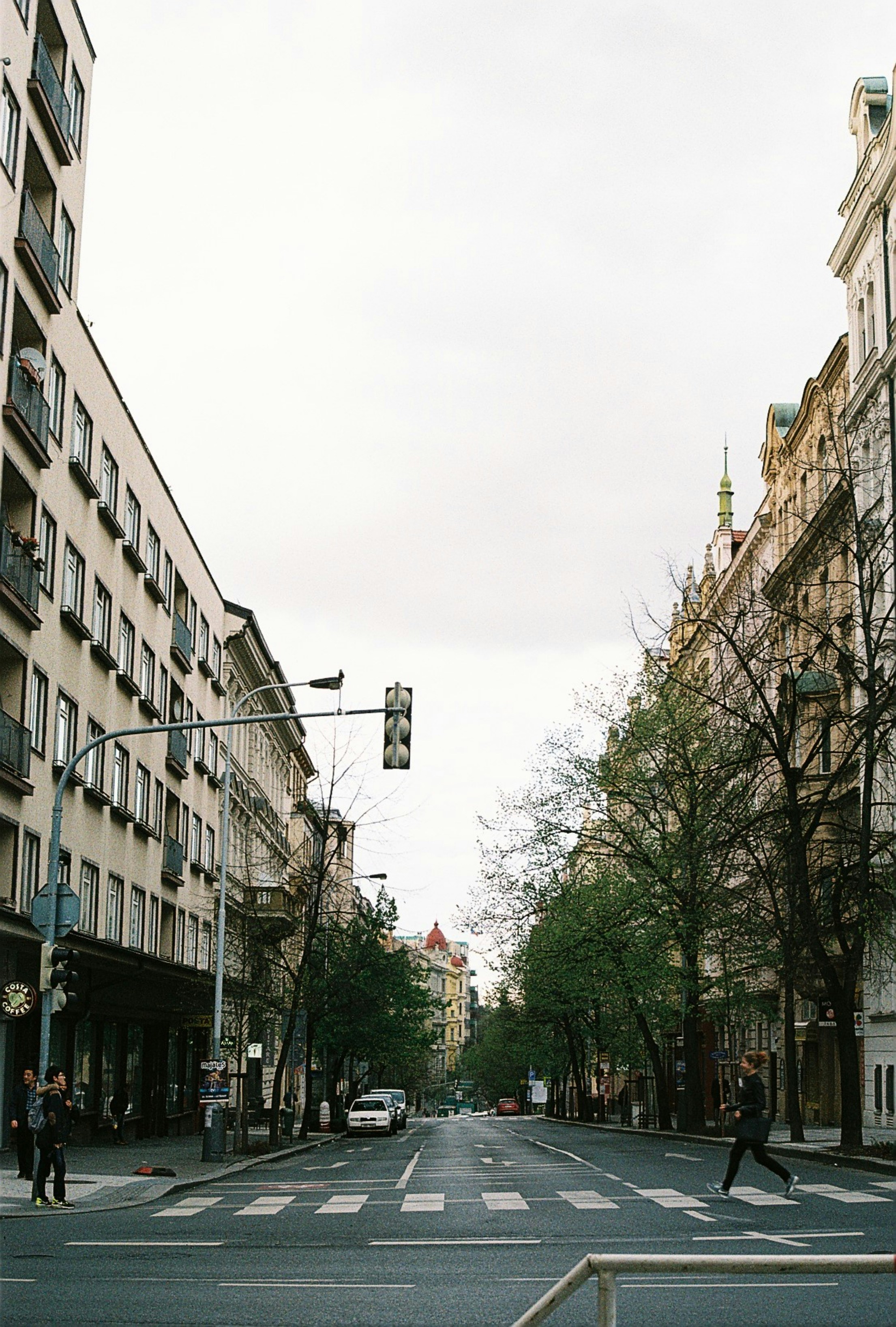 Urban street scene photograph with tall buildings on both sides, a crosswalk, and bare trees along a quiet avenue beneath a pale sky.