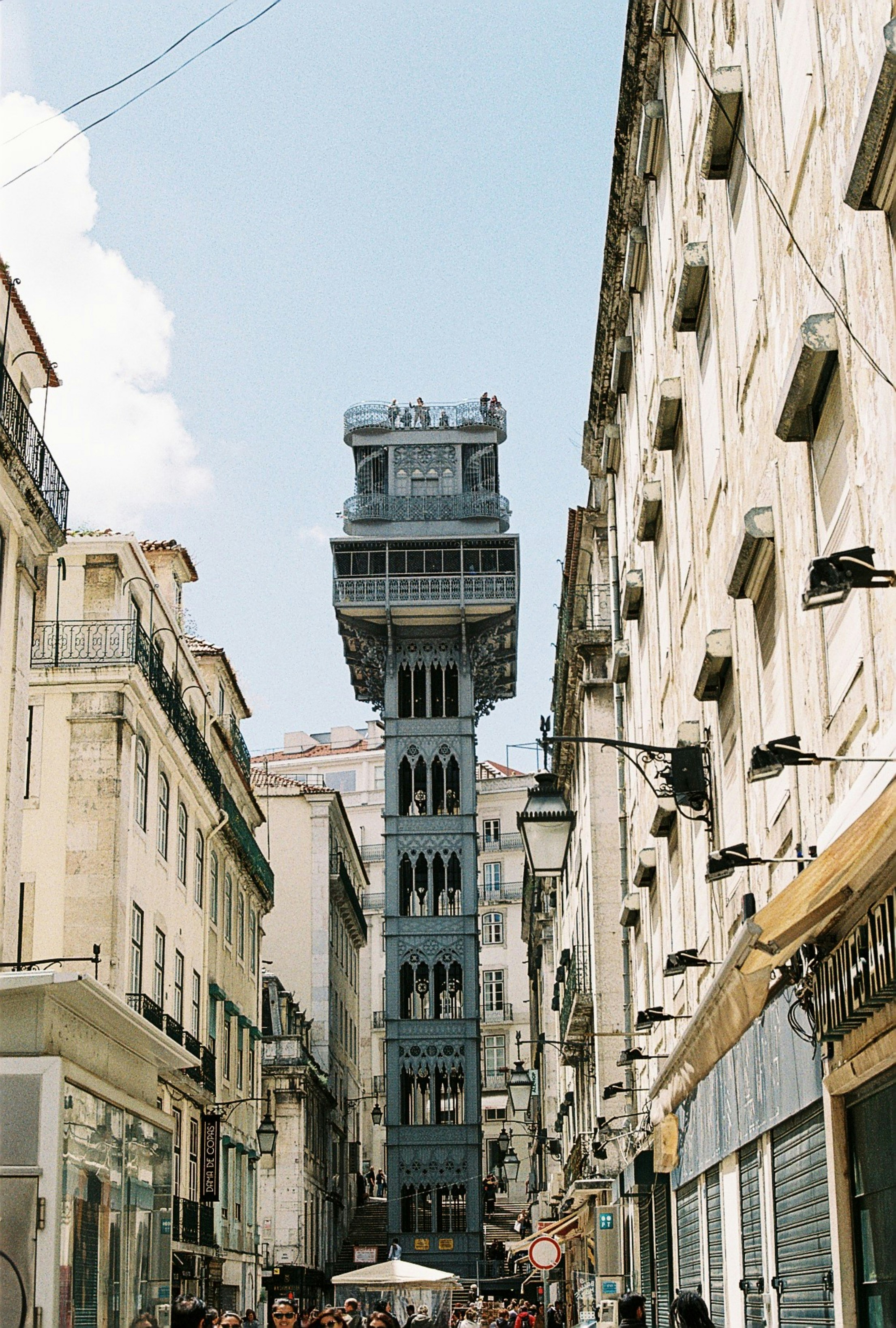 Photograph of Lisbon's Elevador de Santa Justa rising above a sunlit Rua Augusta, flanked by historic buildings and pedestrians.