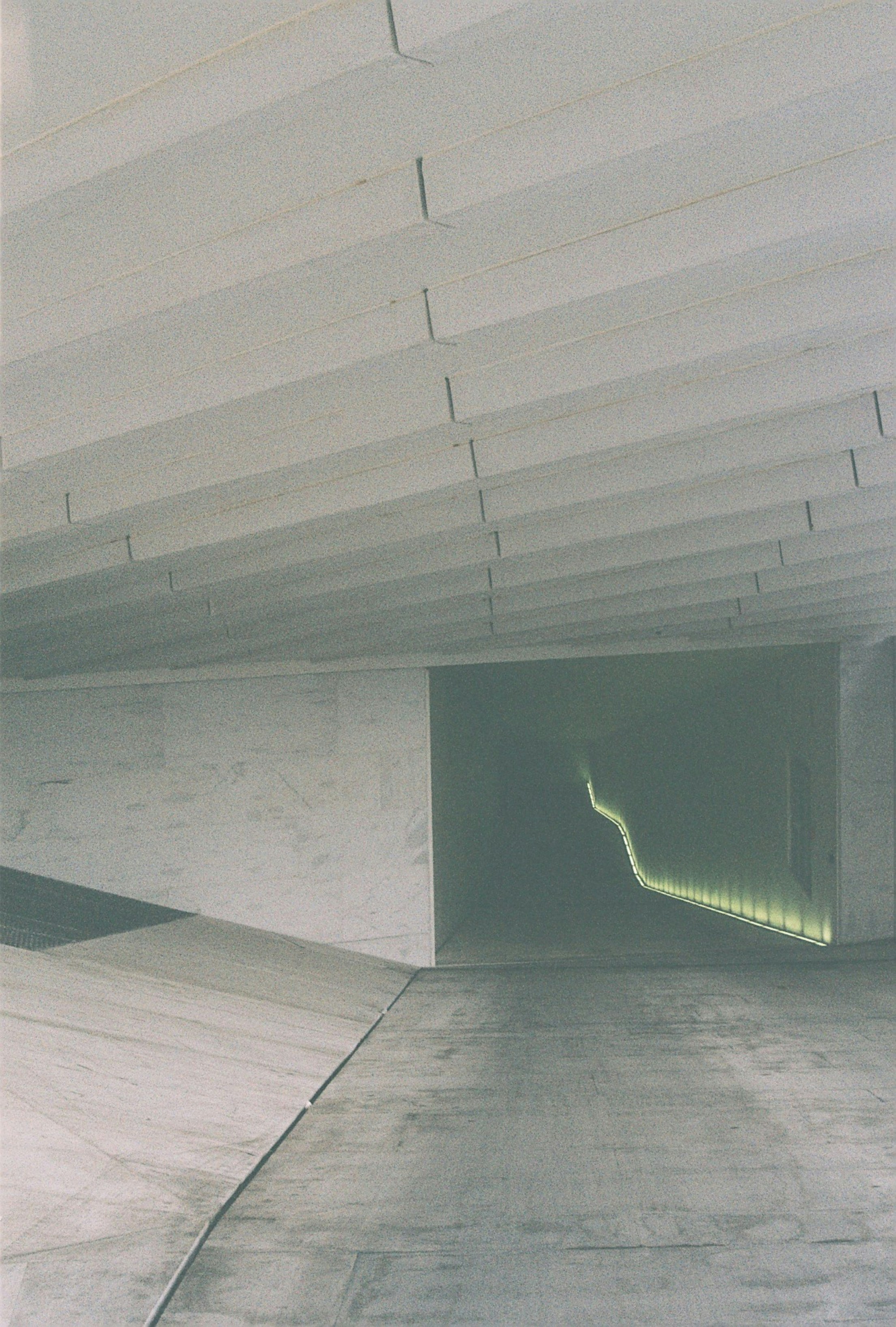 Minimalist architectural photograph of a sloped concrete plaza leading to a dark tunnel, where a neon-green edge traces along the inner wall. The ribbed ceiling and soft desaturated tones emphasize geometry and depth.