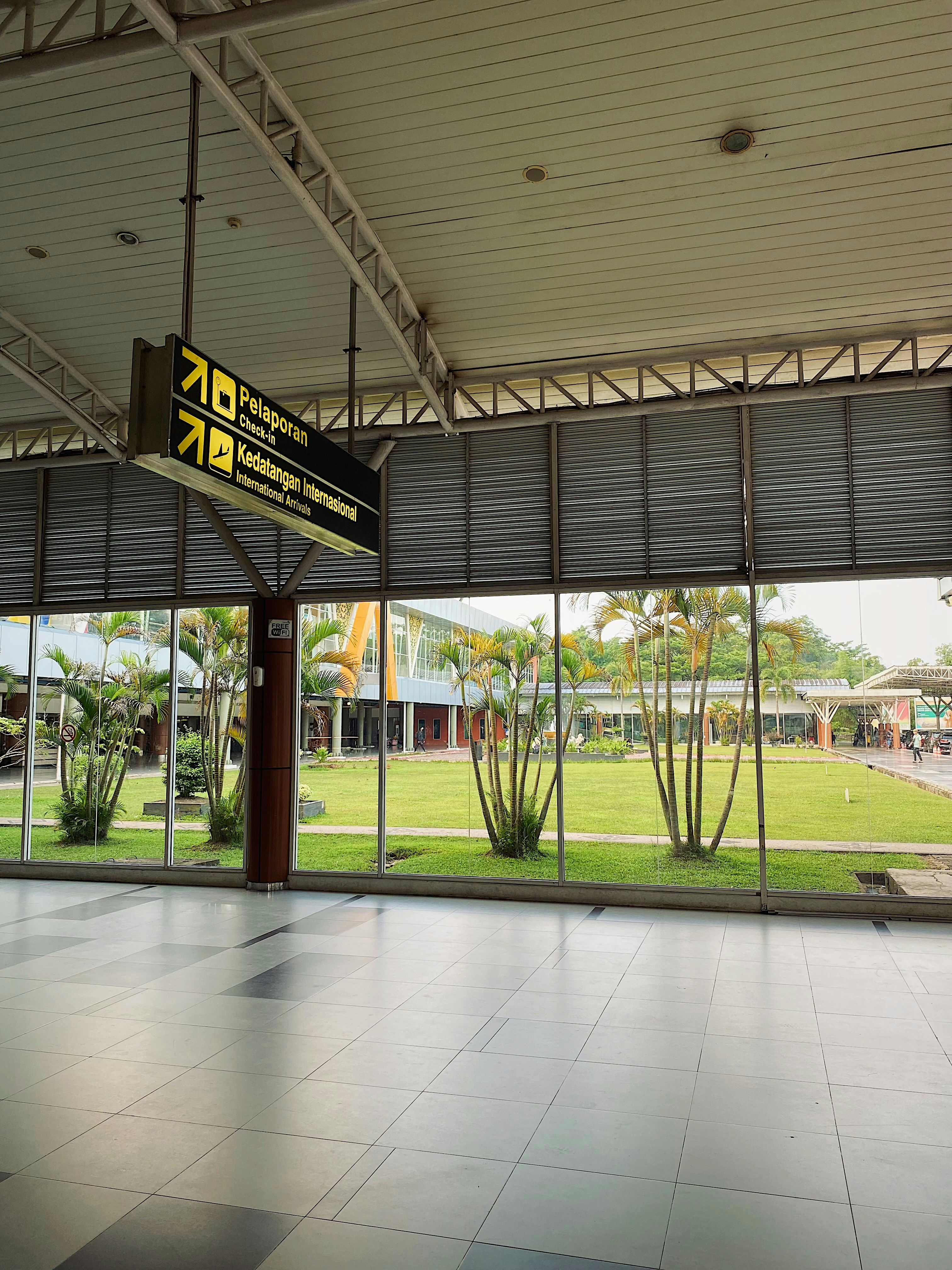 Spacious airport terminal with a directional sign for international arrivals and check-in, overlooking a manicured garden through large windows.