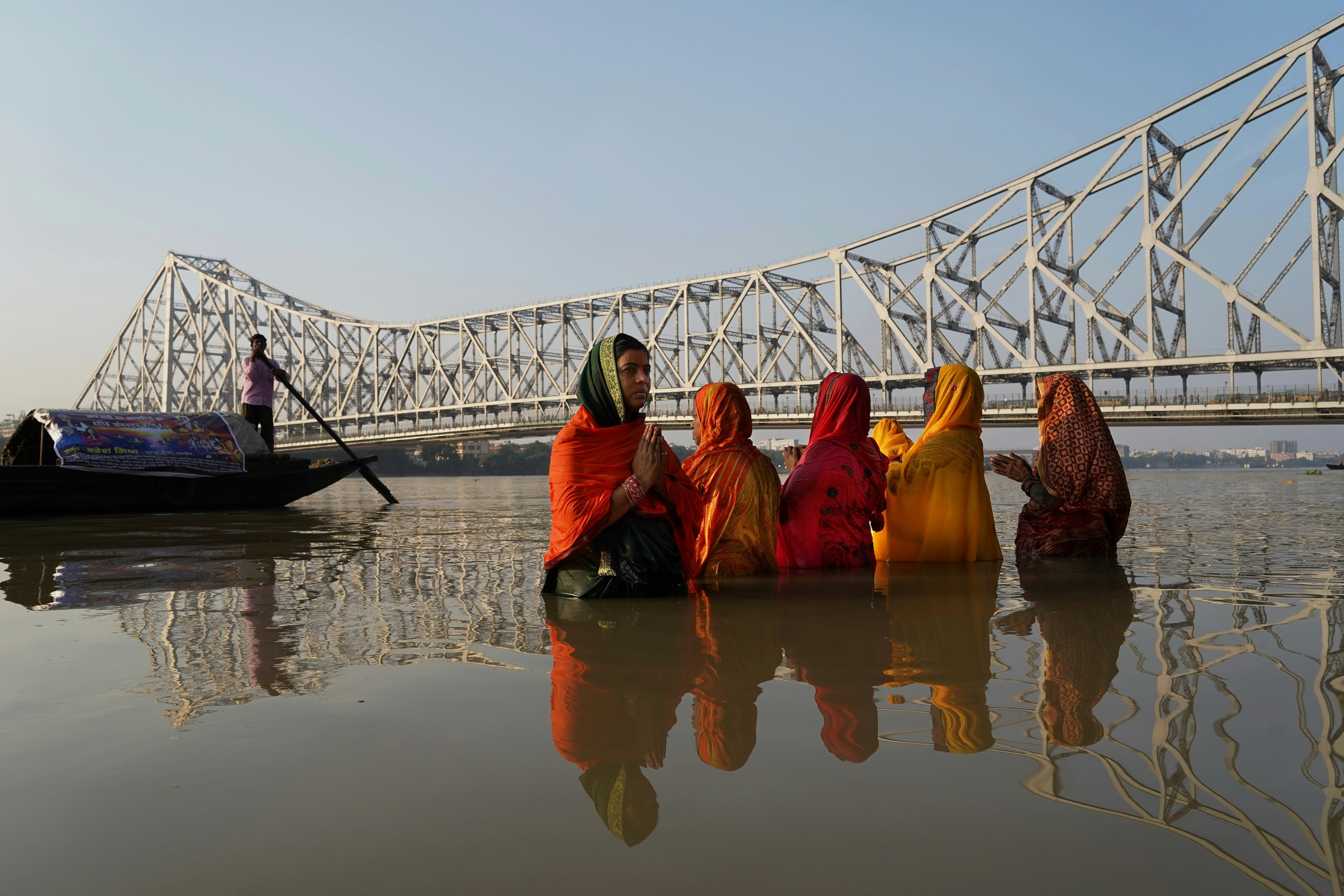 People in colorful attire standing in a river with a large bridge in the background.