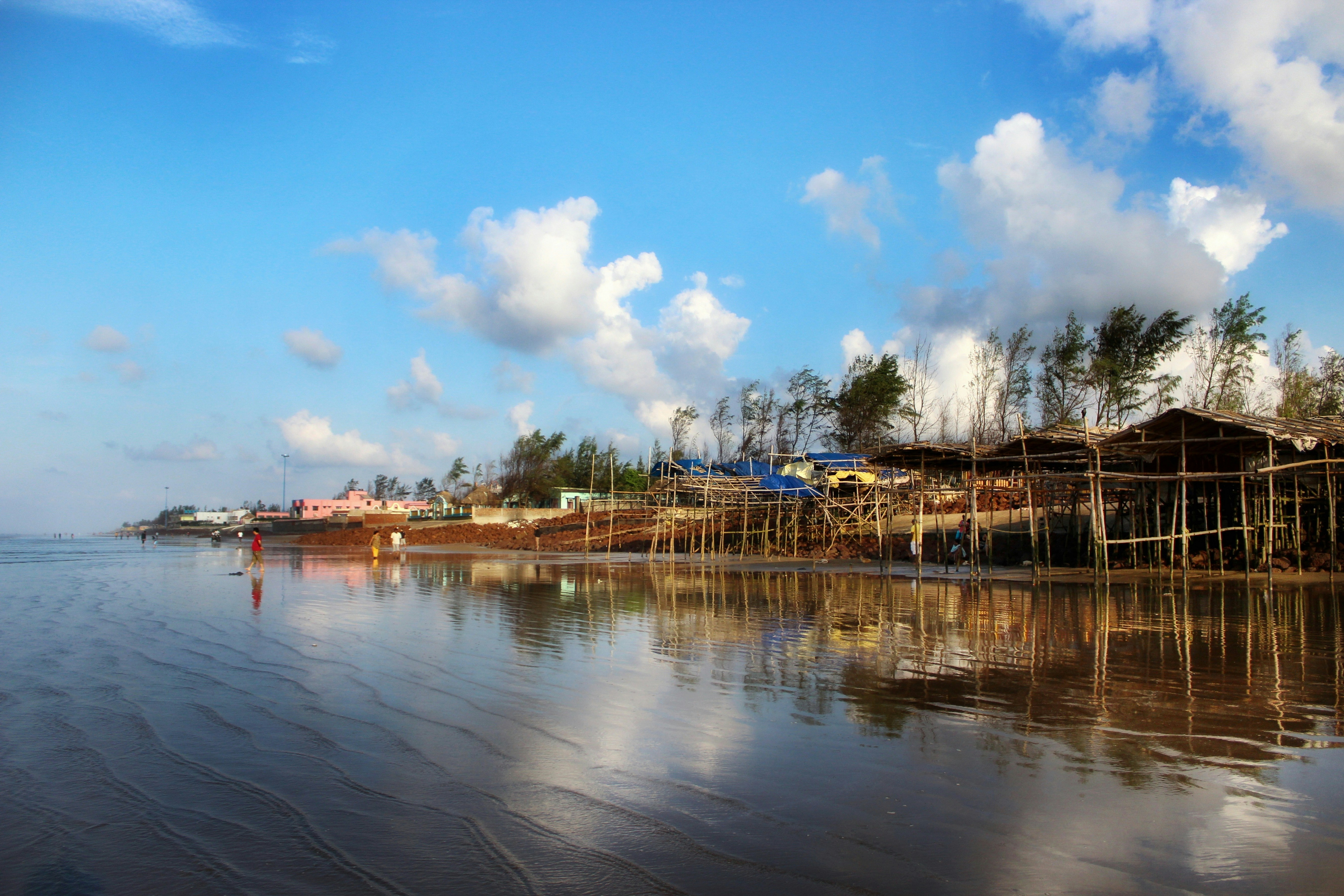 Sandy beach with wooden structures reflecting in calm water under a vibrant blue sky with scattered clouds.